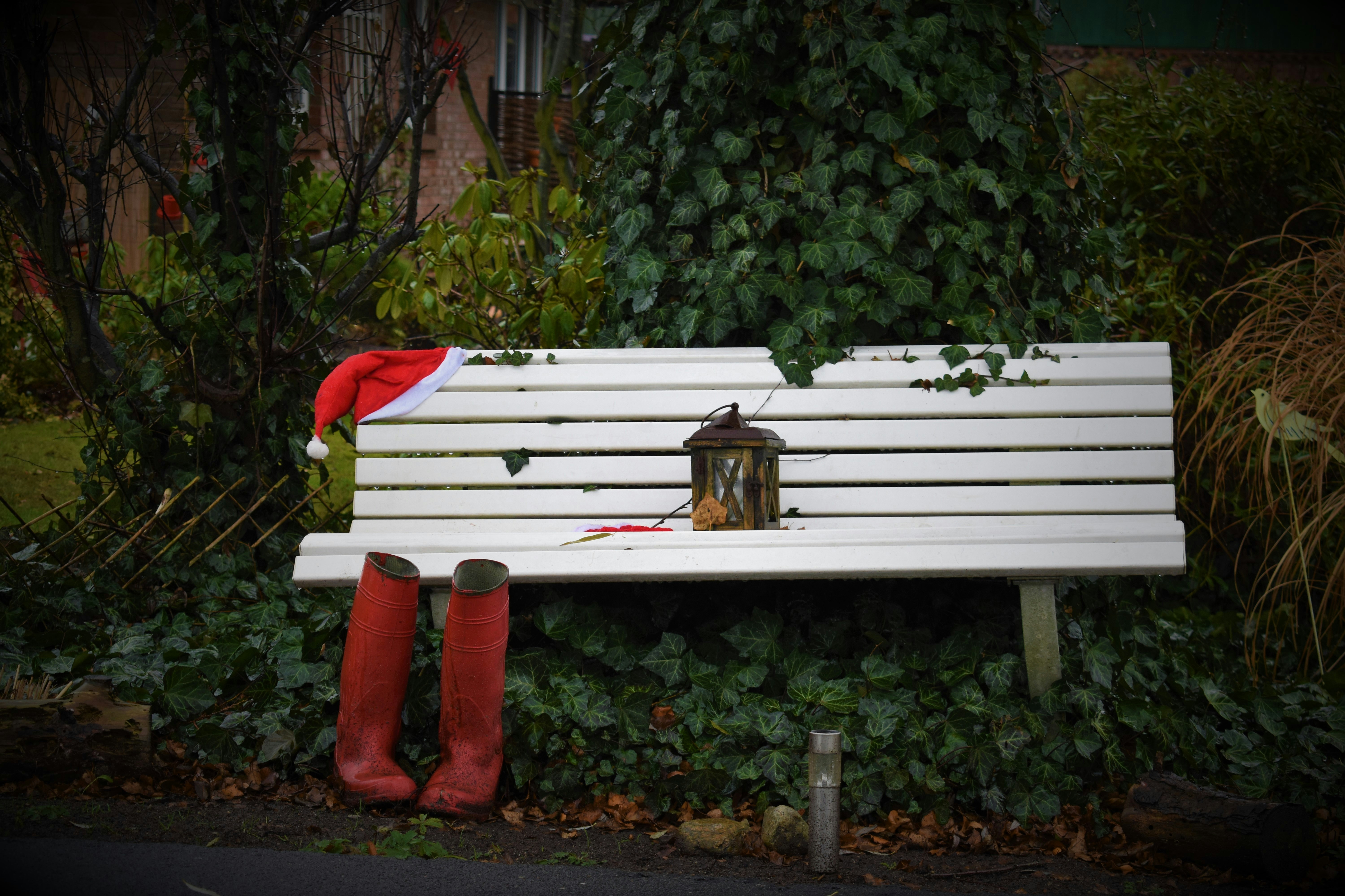 A festive scene featuring a white bench adorned with a Santa hat and a lantern, flanked by vibrant red boots amid lush greenery.