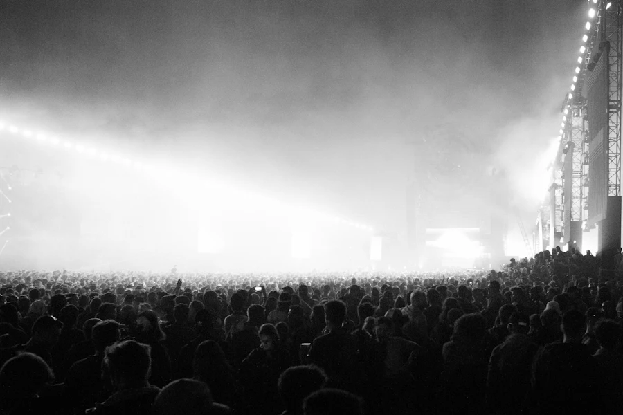 A huge crowd fills an indoor concert or festival venue, silhouetted against an intense wash of white stage light and haze. The stage structures and lighting rigs frame the scene on the right.