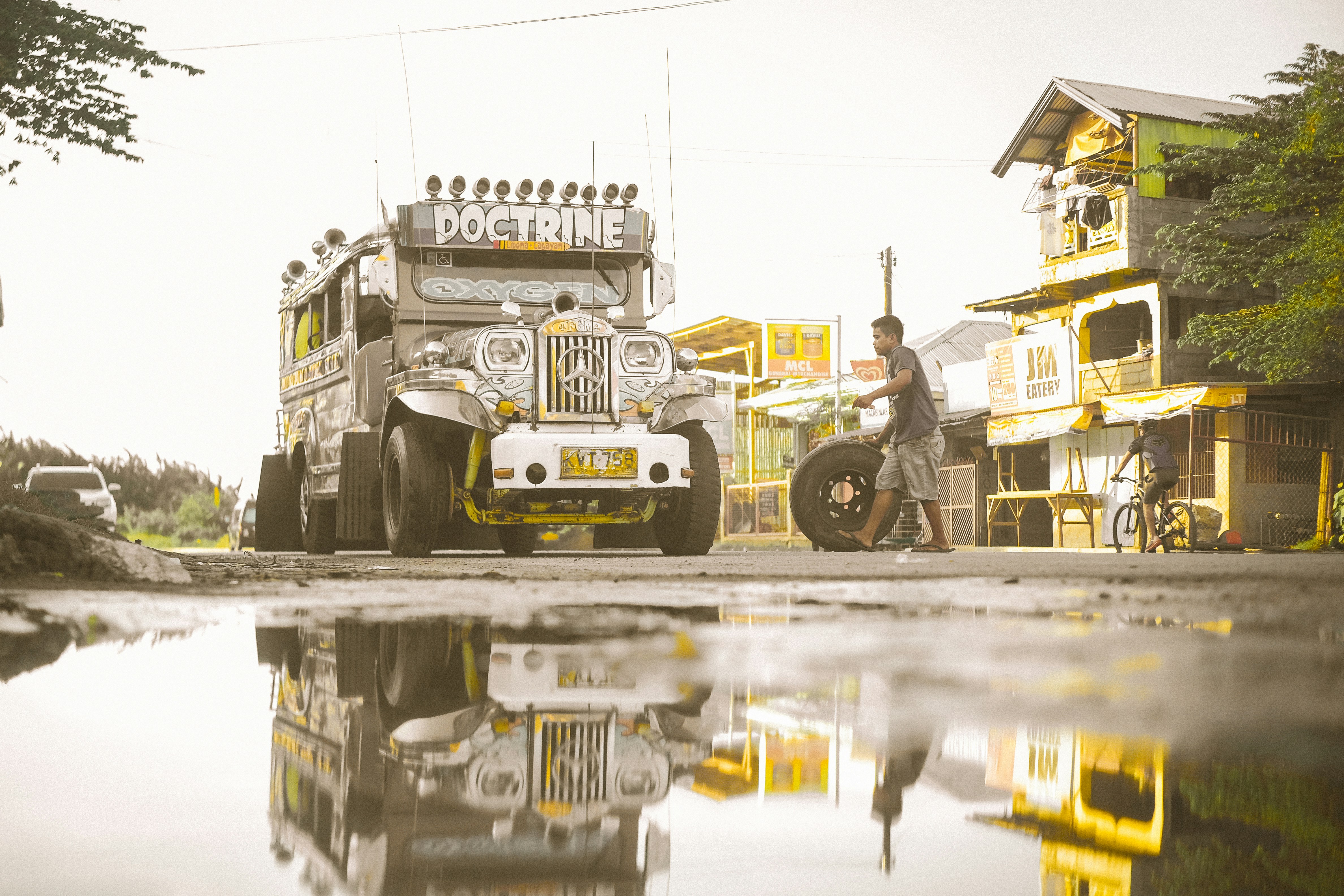 A vibrant jeepney parked by the roadside, reflecting in a puddle, while a man carries a tire nearby. The scene captures the essence of everyday life in a bustling urban environment.