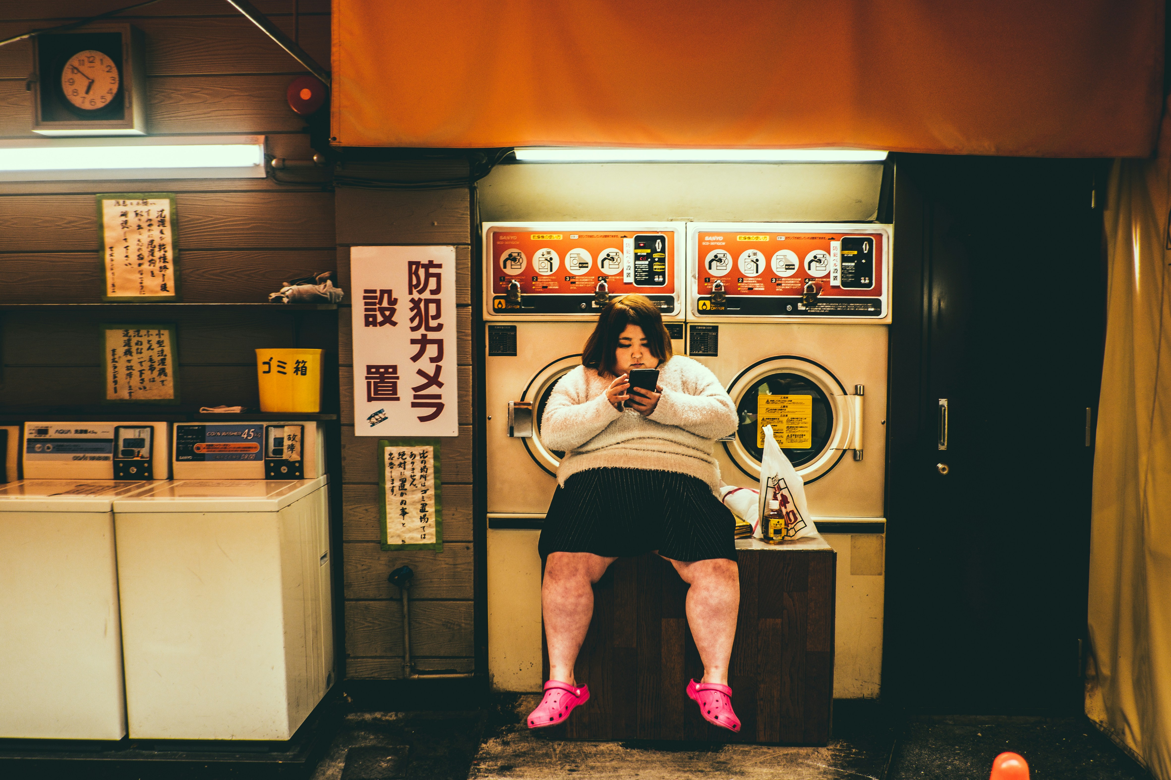 A woman seated on a bench in a laundromat, engrossed in her phone while surrounded by washing machines and signs. The scene captures a blend of everyday life and personal reflection.