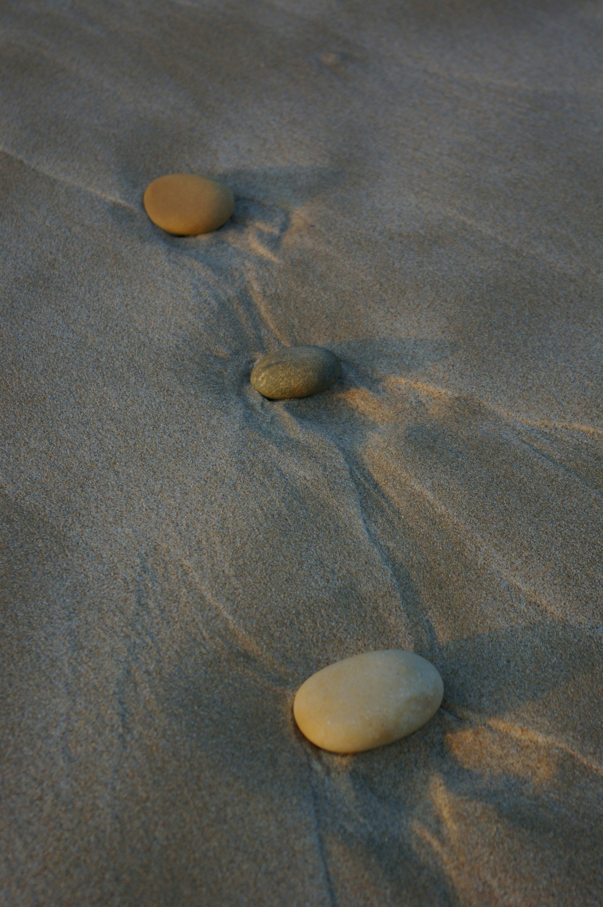 Three smooth pebbles rest on sunlit sand, their rounded forms echoed by wind-carved grooves.