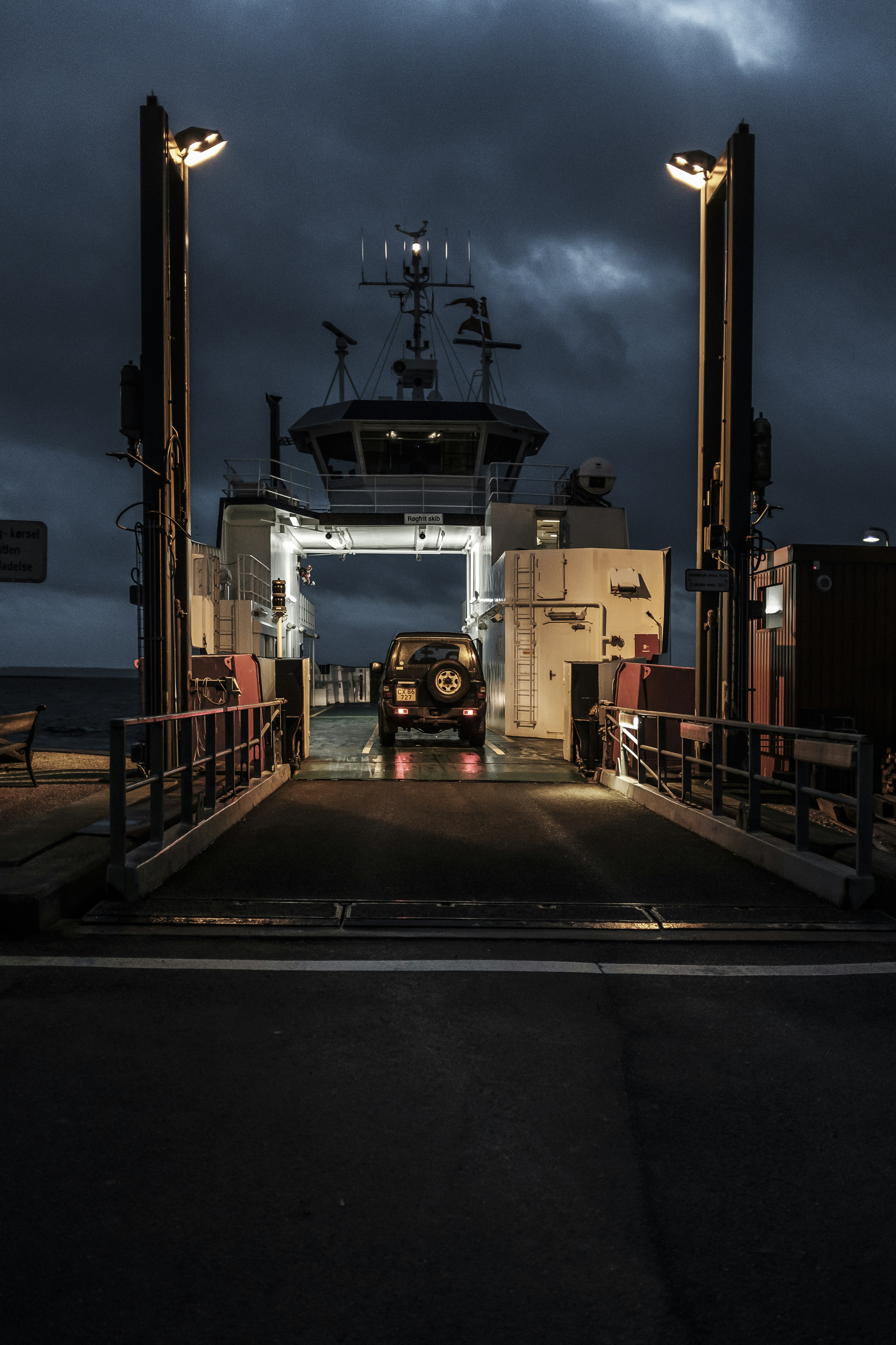 A vehicle poised to embark onto a ferry under a dramatic sky, illuminated by the vessel's bright lights. The scene captures the essence of travel and adventure.