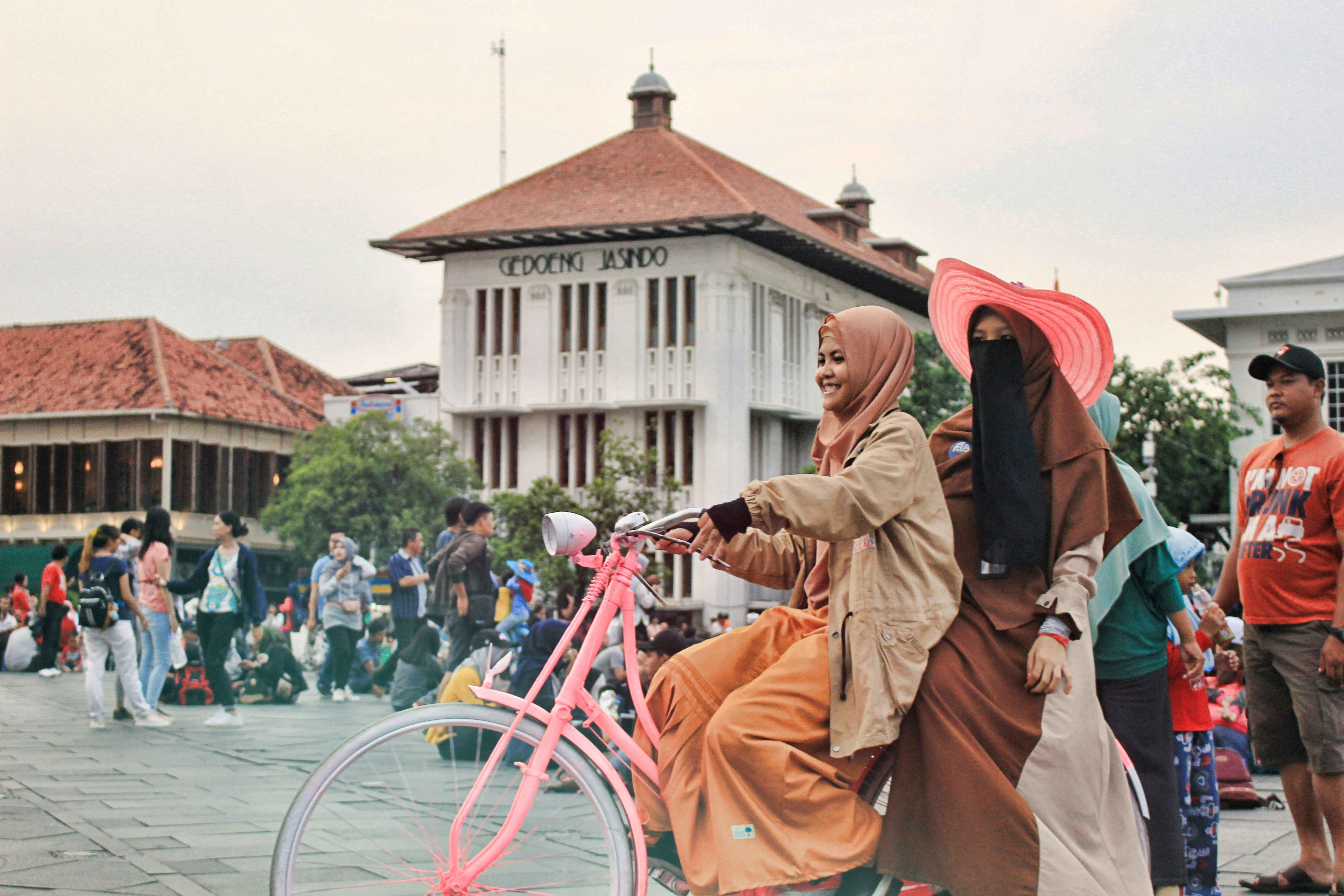 Two women ride a bright pink bicycle past a bustling crowd near a historic building in Jakarta.