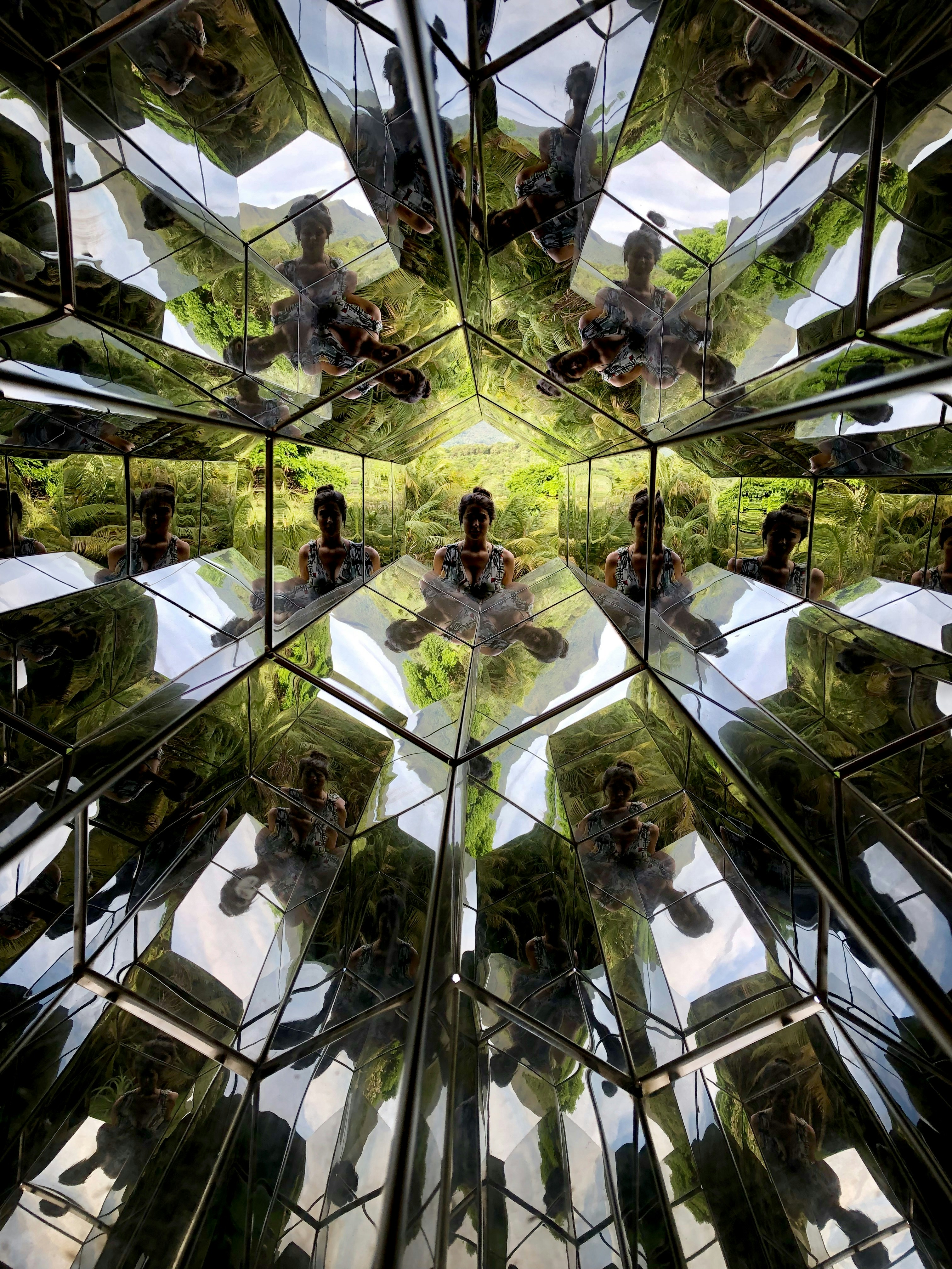 Mirrored installation capturing multiple reflections of visitors amidst lush greenery. The intricate design creates a kaleidoscopic effect.