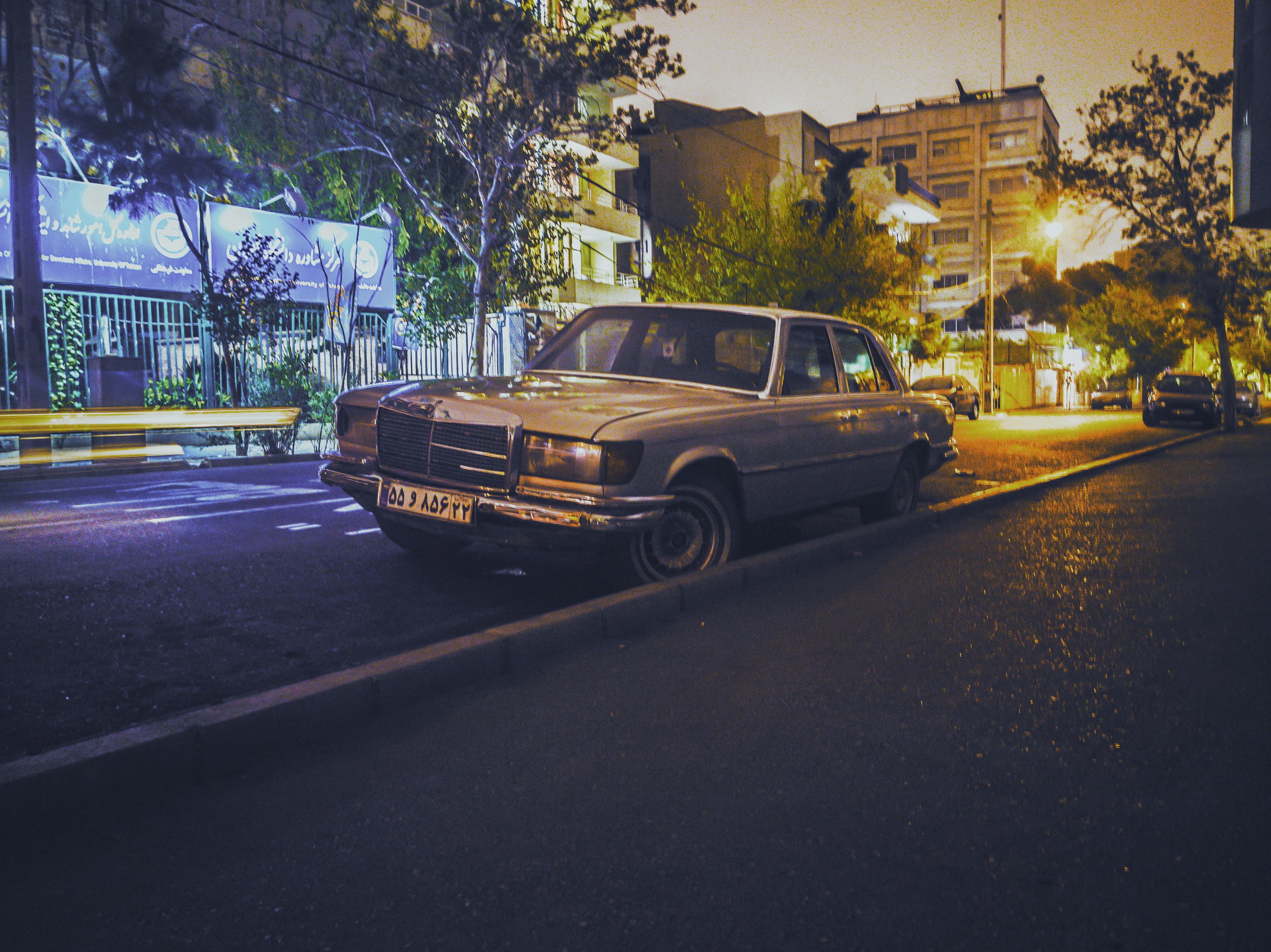 Classic silver sedan parked on a quiet urban street, illuminated by warm streetlights and surrounded by greenery.