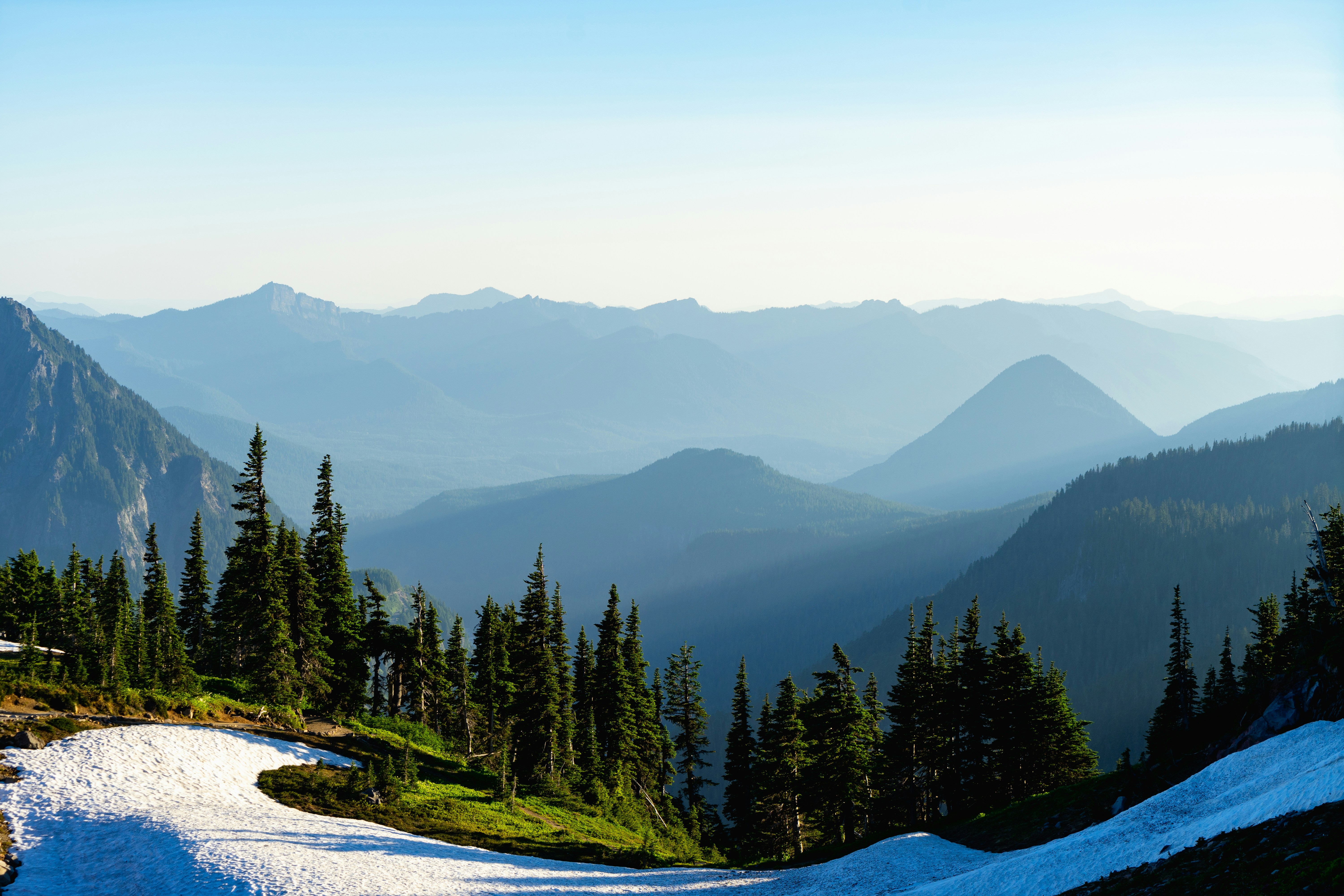 Snow patches and evergreen trees overlooking layered mountain ranges under a clear blue sky.