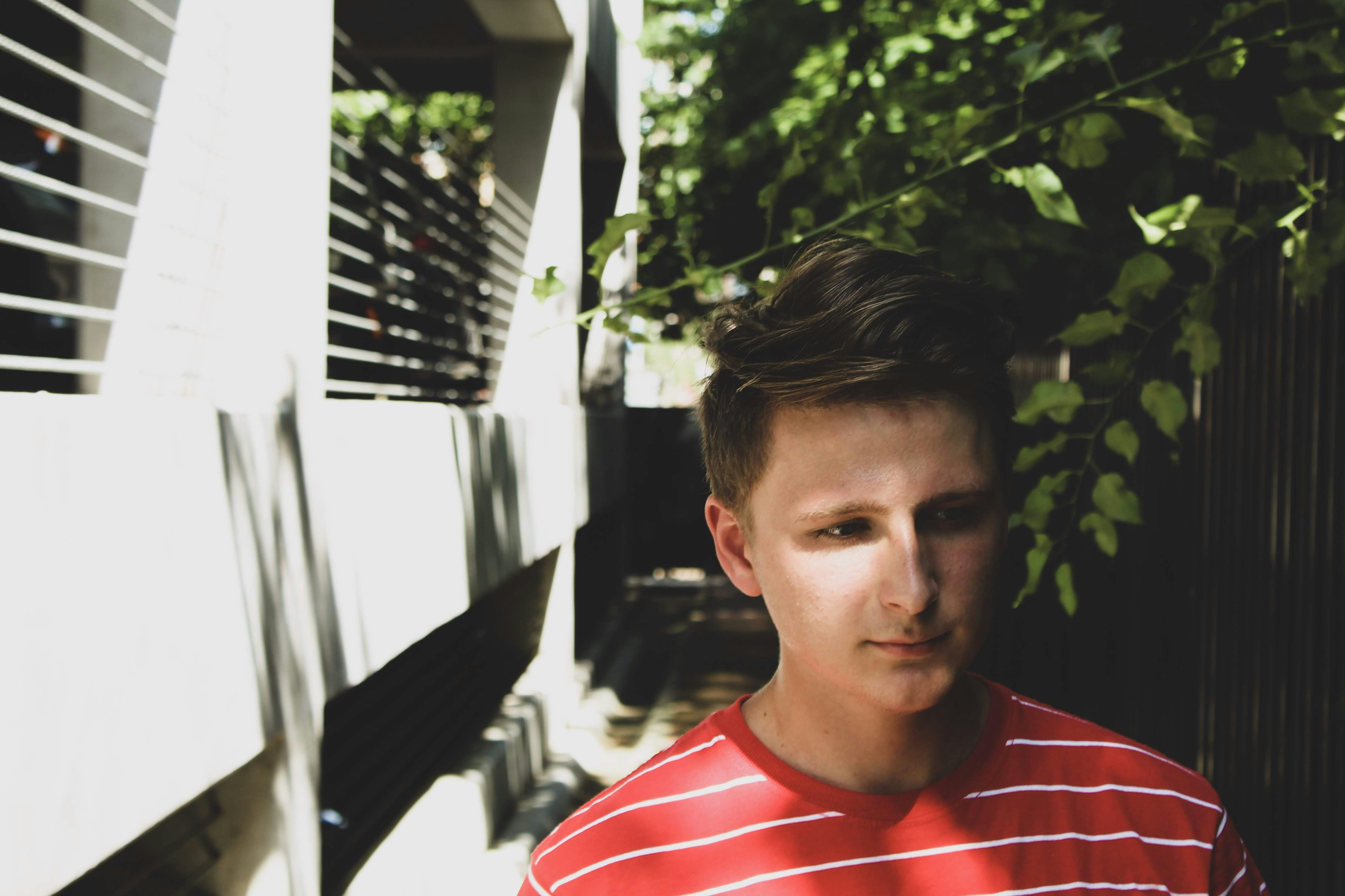 Person in a red striped shirt stands in a shadowed alley with dappled sunlight filtering through leaves.