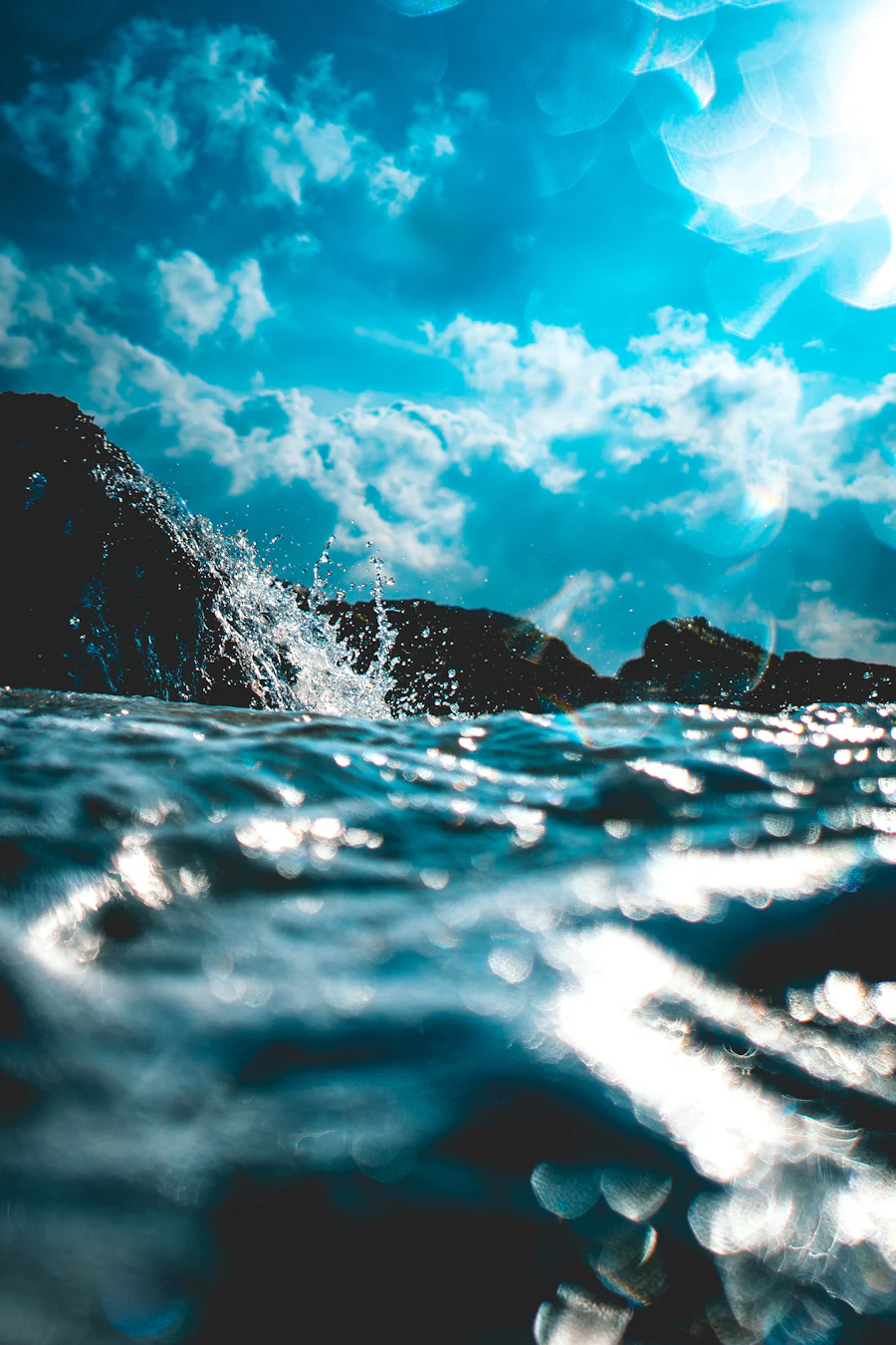 Waves crash against jagged rocks under a bright blue sky, with water droplets catching light in the foreground. A dynamic seascape captures motion and texture of spray and ripples.