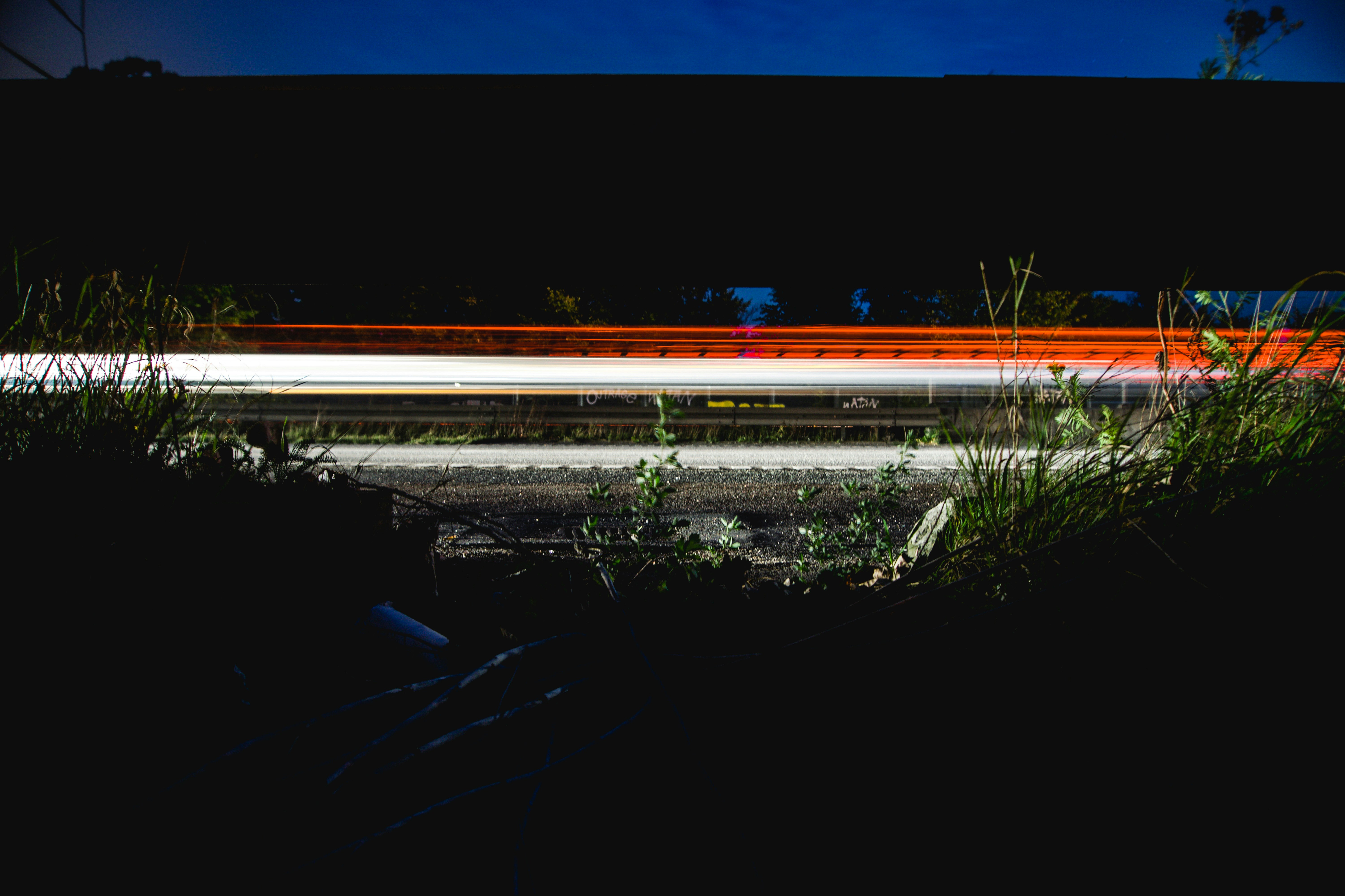 Long exposure capture of vehicle lights streaking along a road, framed by grass and foliage, under a twilight sky.