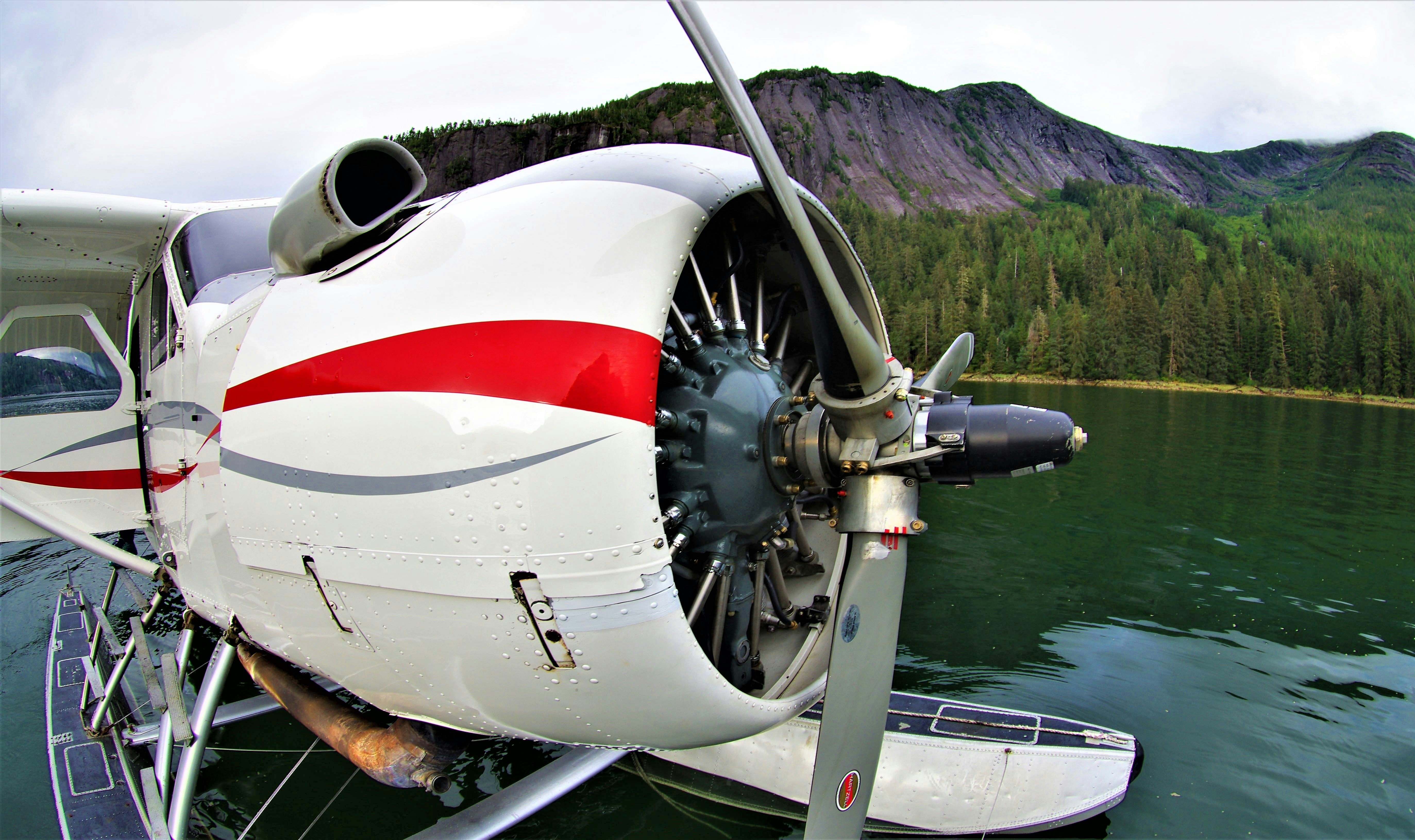 Close-up of a seaplane's engine and propeller reflecting on calm waters, with a backdrop of lush mountains and trees.