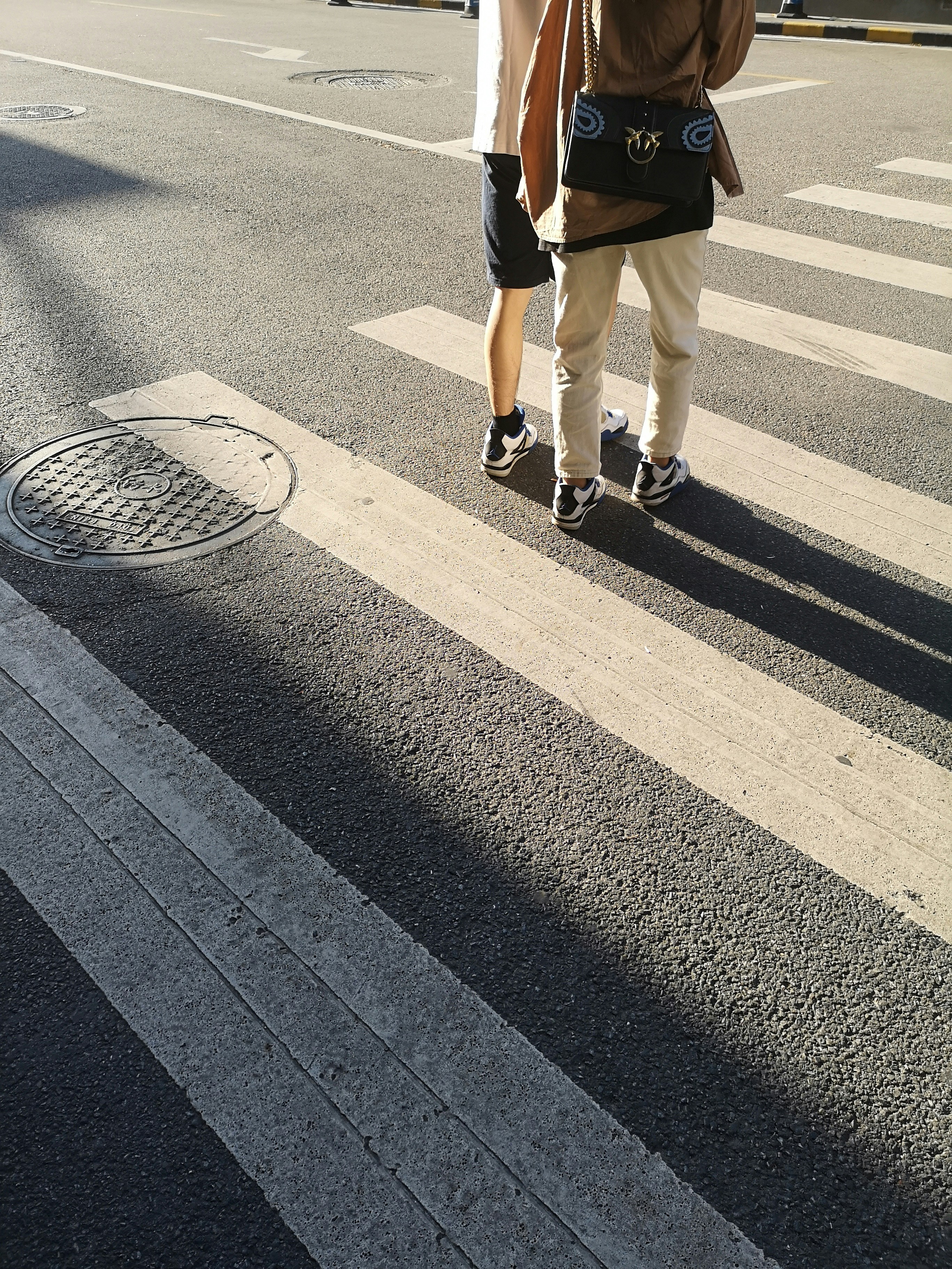 Two individuals standing together at a crosswalk, casting long shadows on the asphalt. Their footwear and casual attire reflect a relaxed urban lifestyle.