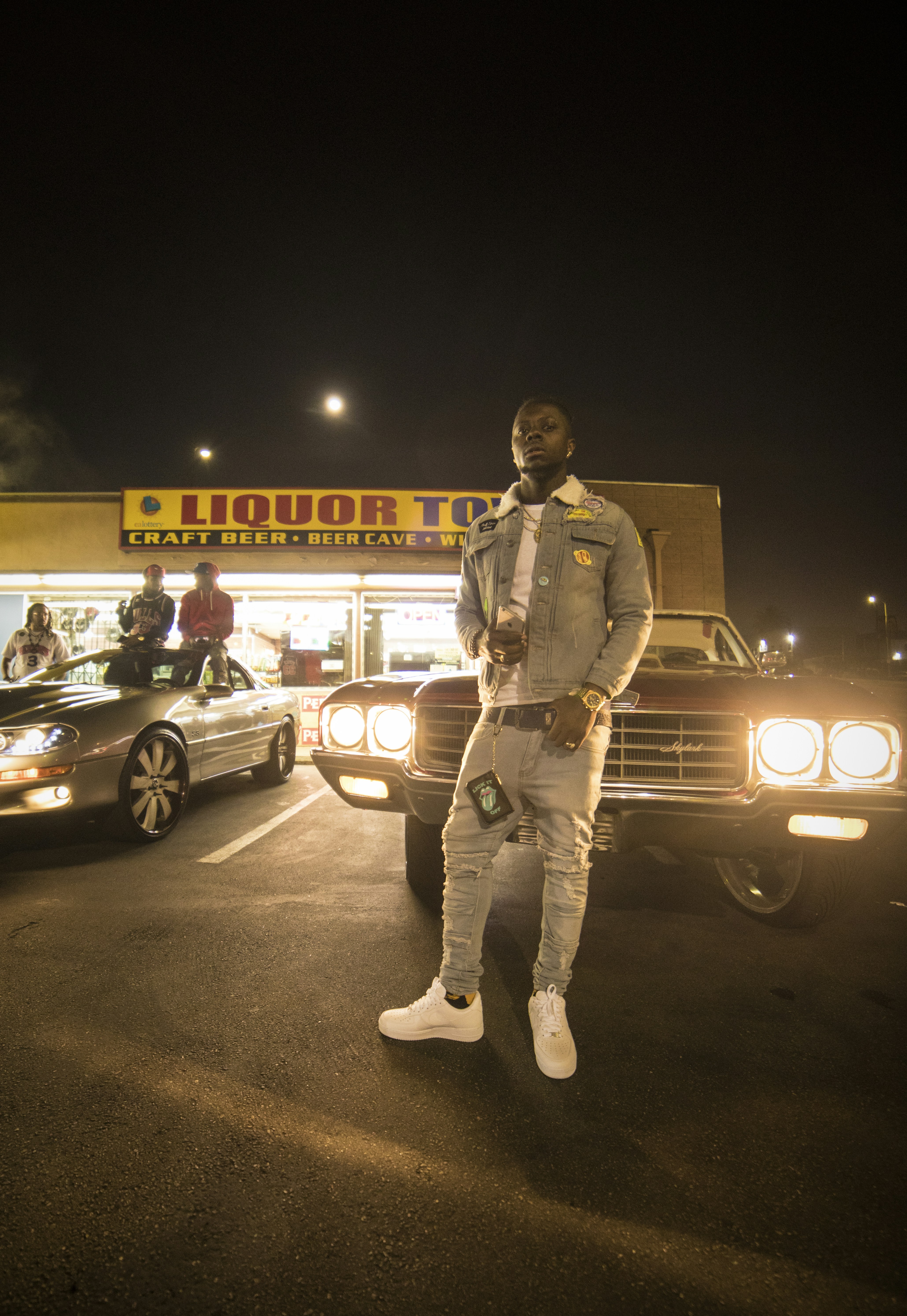 Man in front of vintage car outside a well-lit liquor store at night.