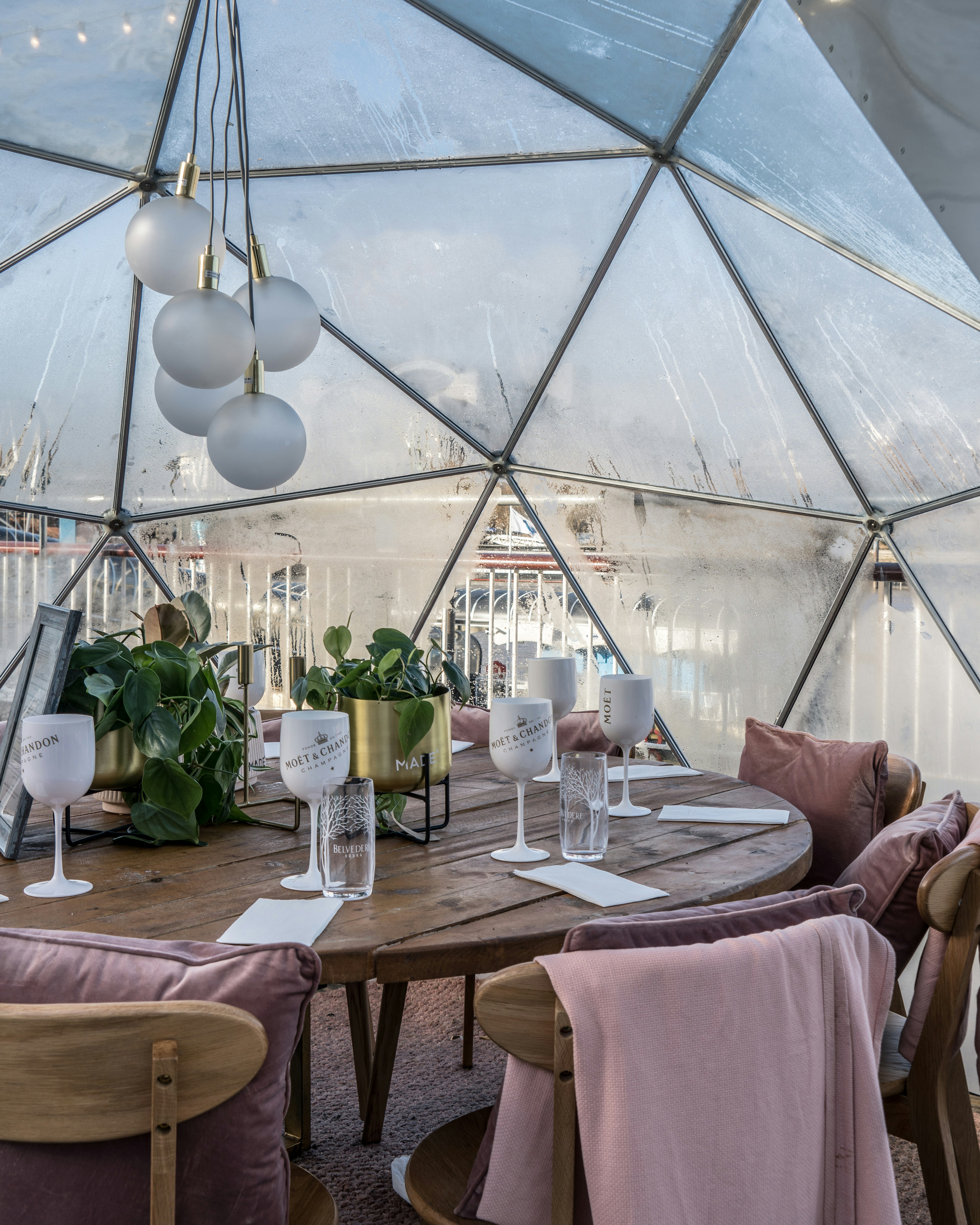 Wooden table set with wine glasses inside a glass igloo with lush greenery and soft pink cushions.