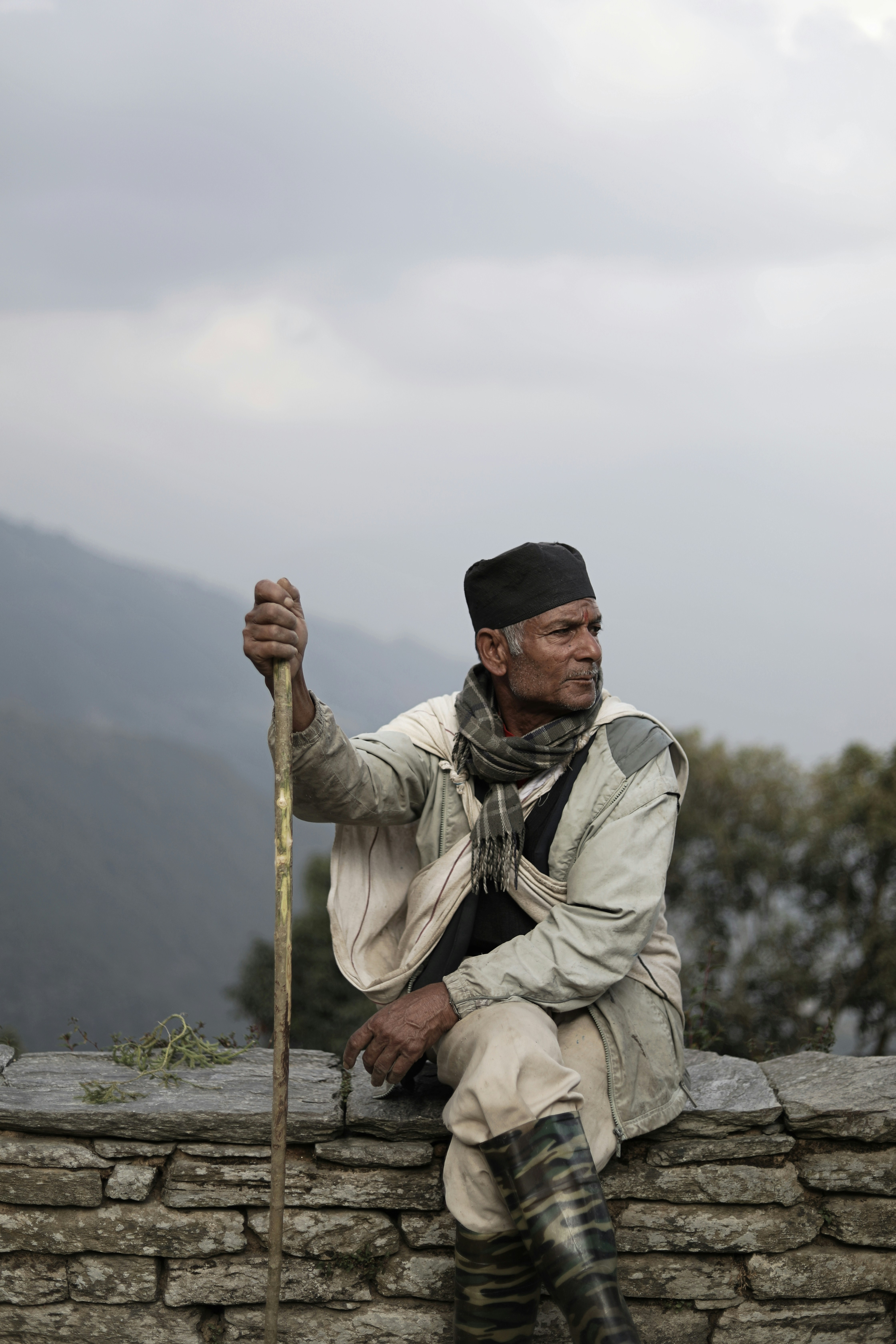 Elderly man seated on a stone wall, gazing thoughtfully into the distance with a walking stick in hand. The misty mountains create a serene backdrop.