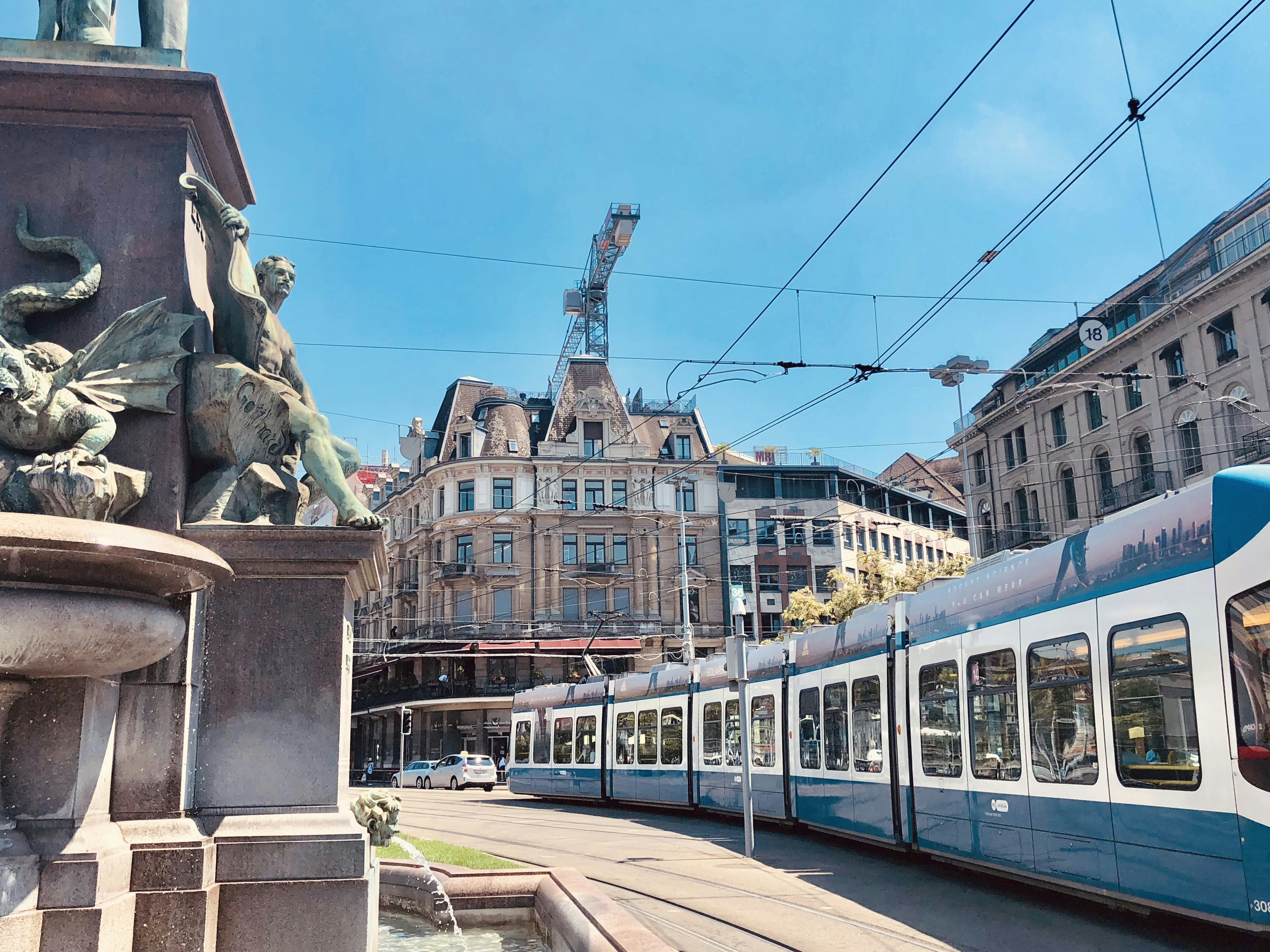 Tram passing a historic statue in a vibrant city square under a clear blue sky.