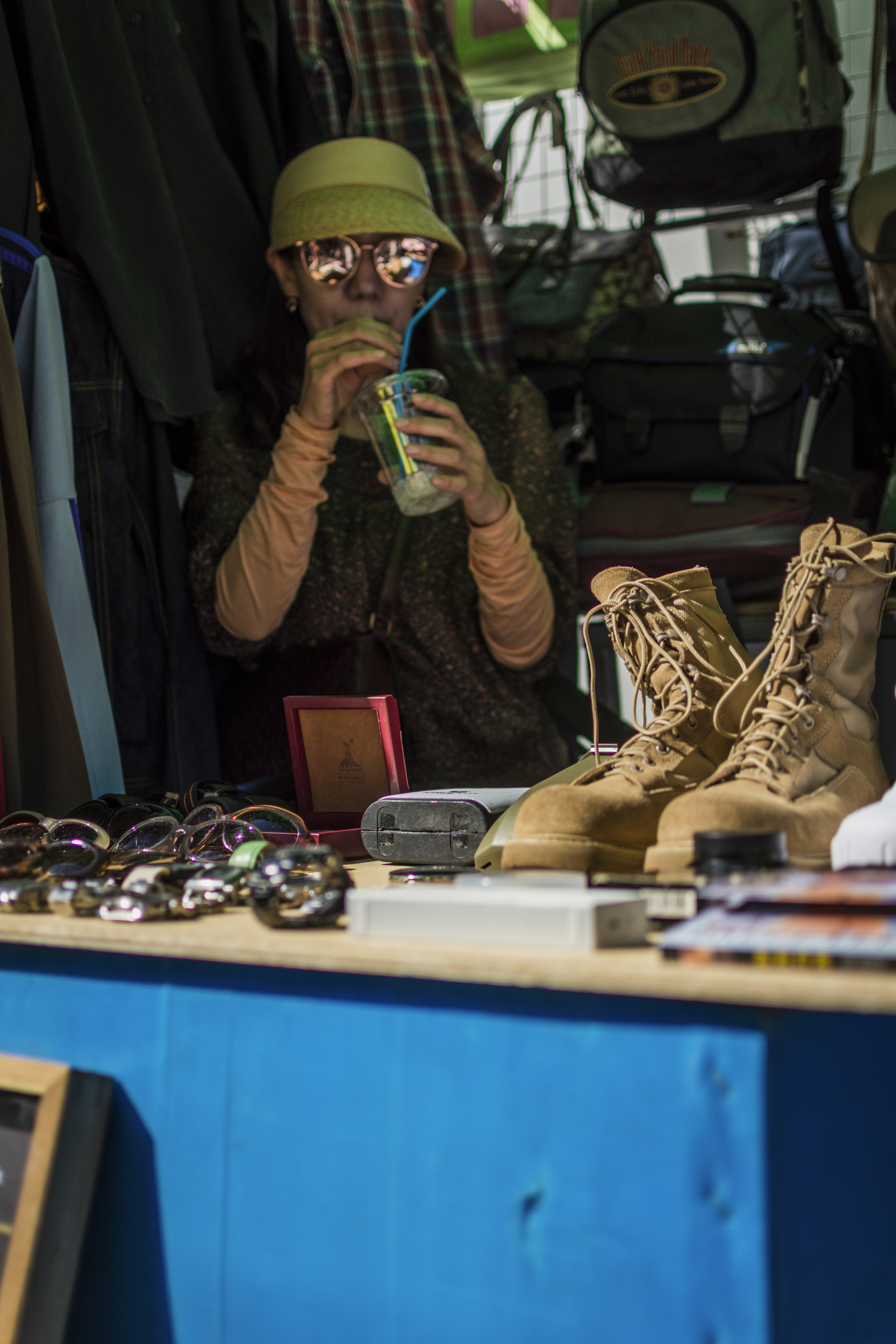 A vendor sipping a drink amidst an array of sunglasses and boots in a vibrant market stall.