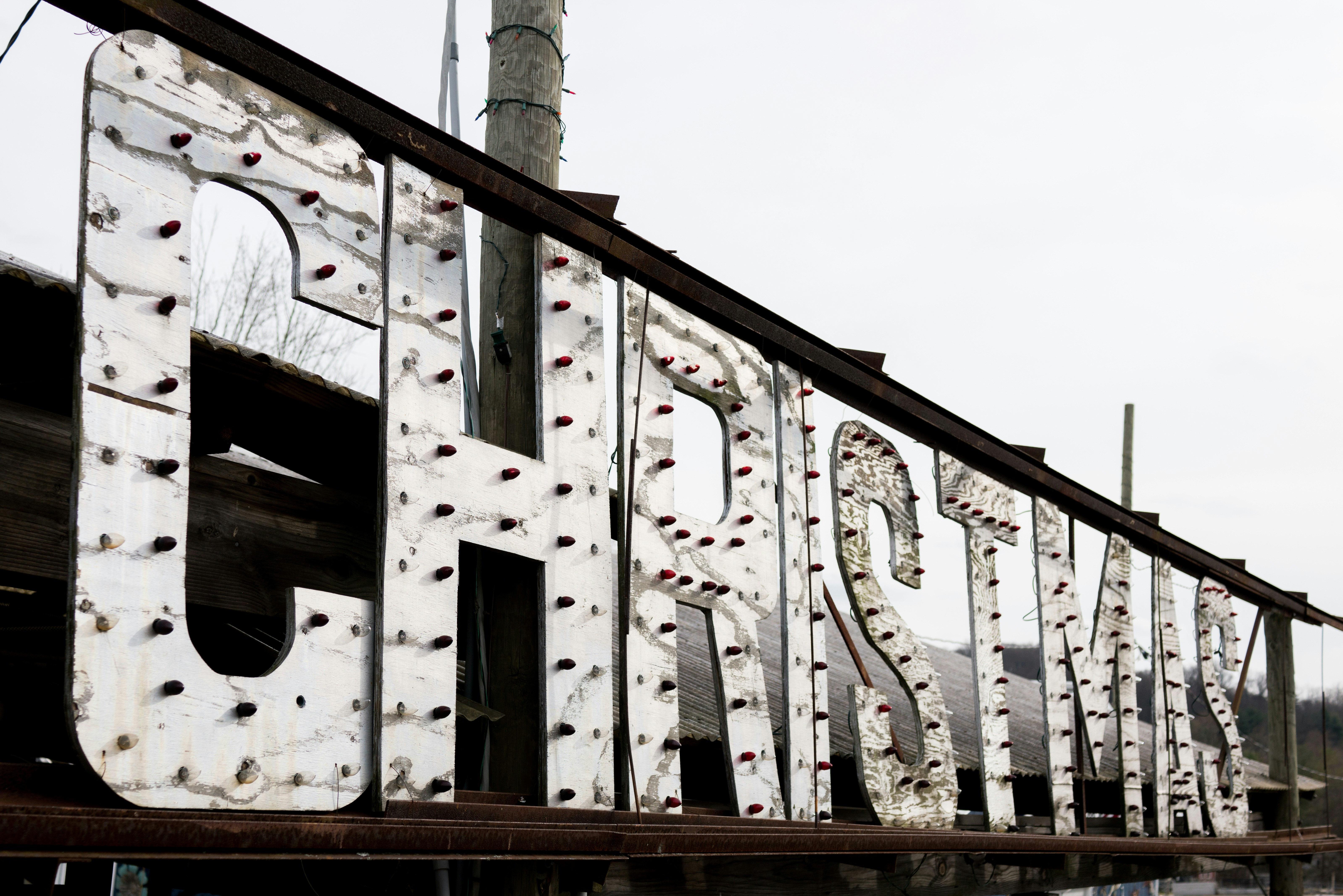 Large weathered metal sign with perforated, bullet-holed letters spelling CHRIST stretches along a rusted frame against a pale sky.