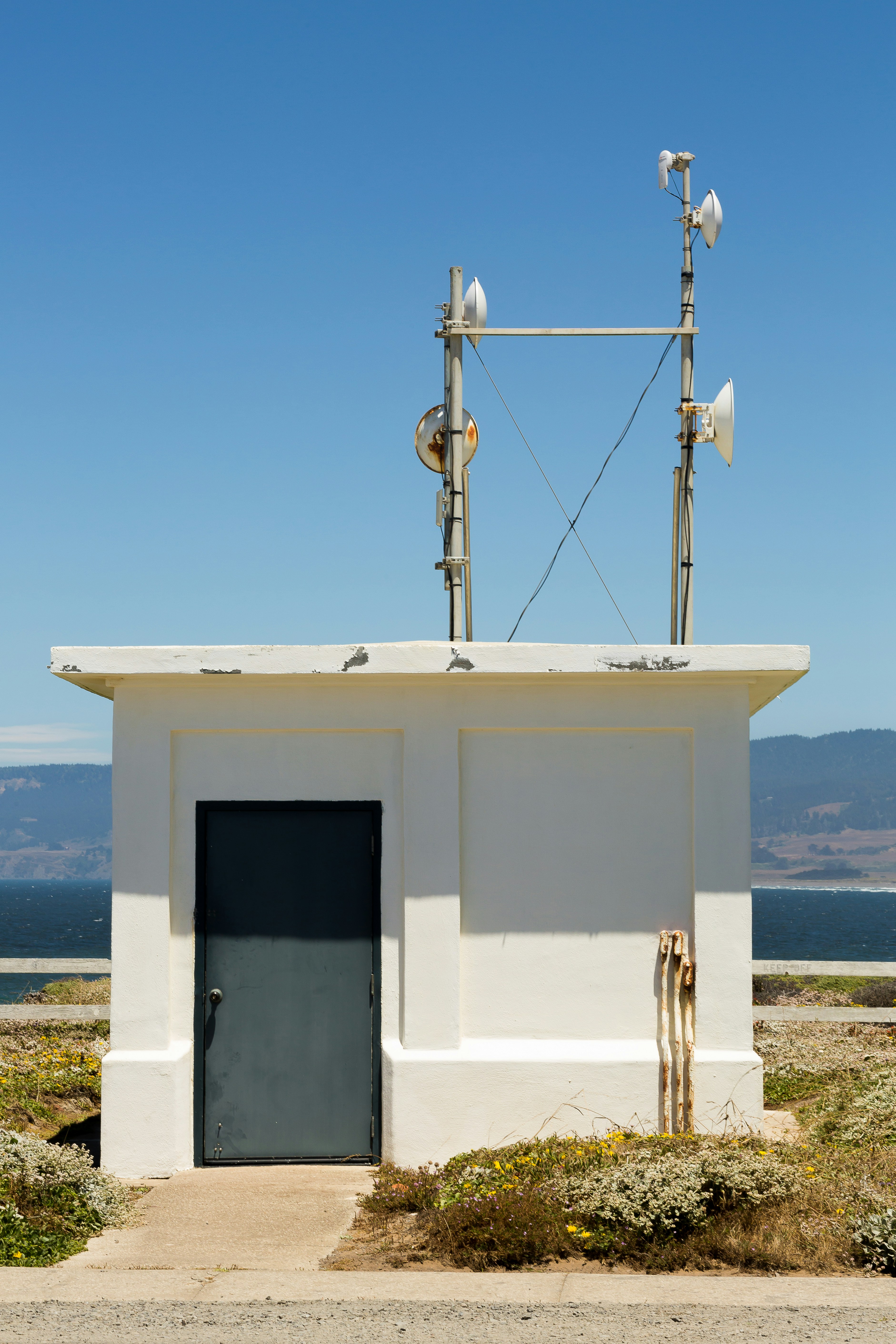 A small, white communication building with antennas and speakers atop, set against a clear blue sky and distant hills. The structure stands as a quiet observer of the surrounding landscape.
