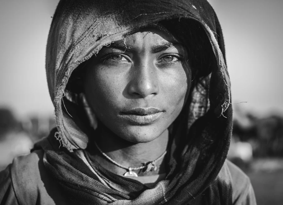 Close-up black-and-white portrait of a woman wearing a hooded scarf, looking directly at the camera with a serious expression. Soft background blur keeps focus on her face and eyes.