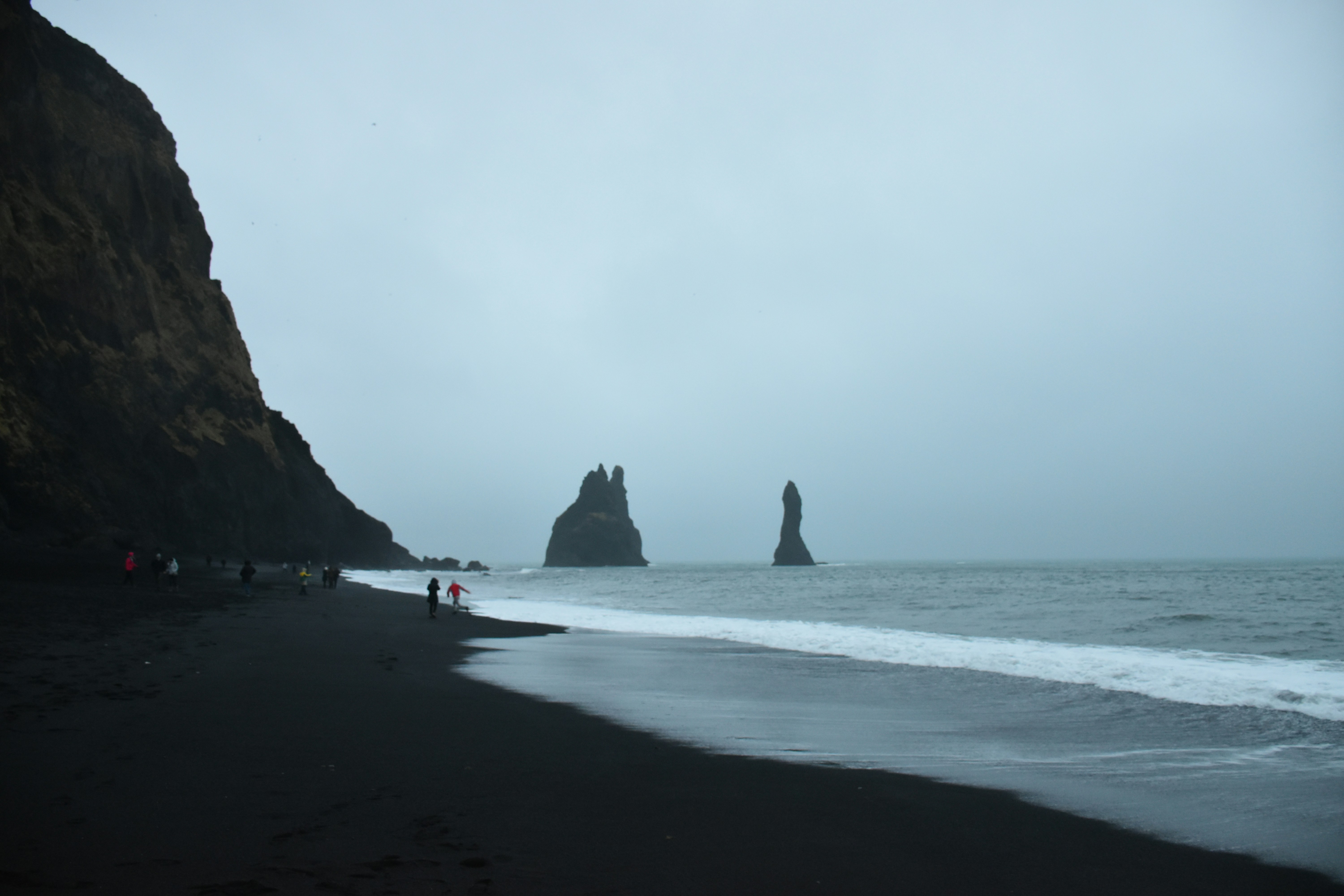People walking along a black sand beach with towering sea stacks in the distance on a cloudy day.