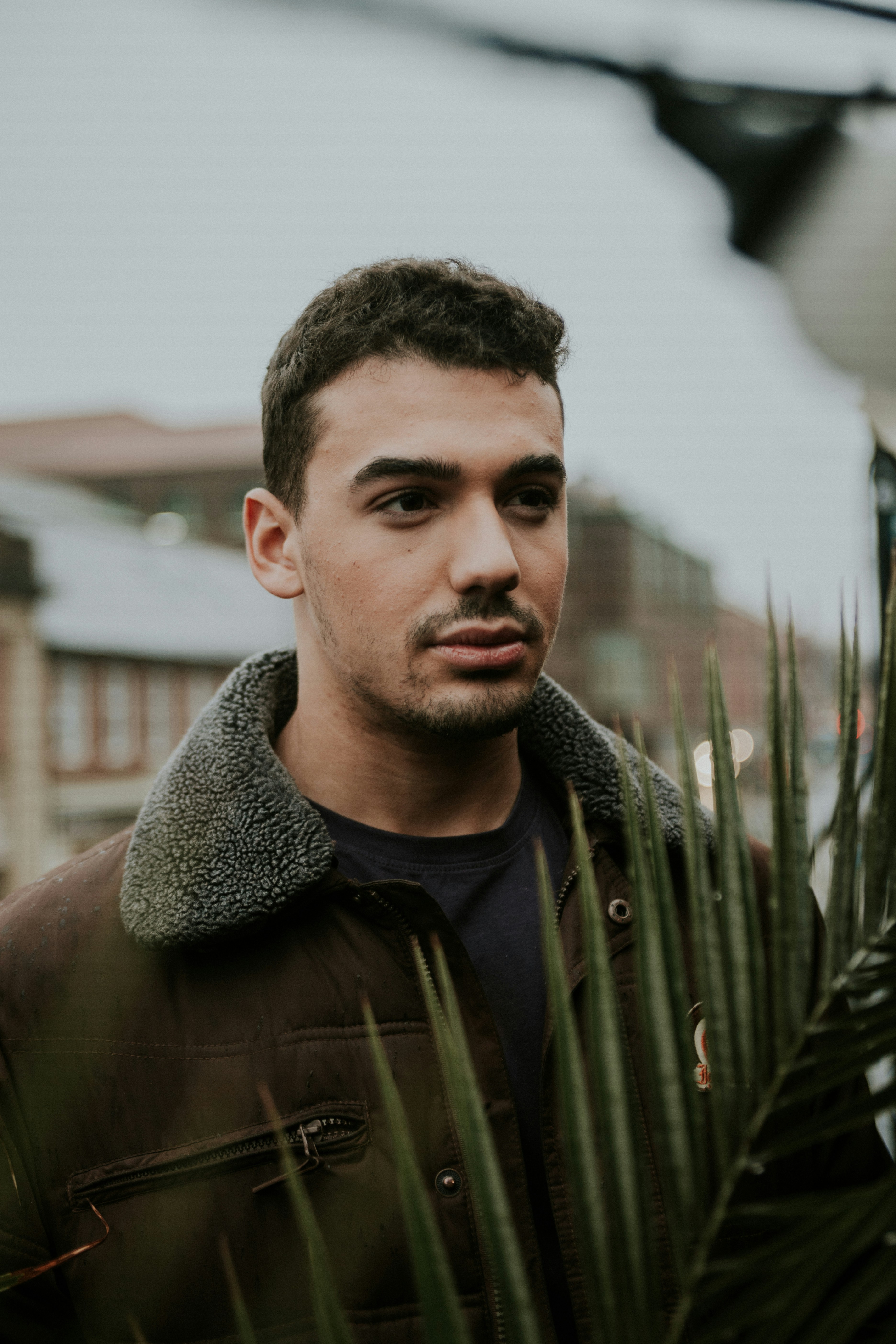 Young man gazing thoughtfully amidst greenery in an urban setting. The contrast between nature and city life is highlighted.