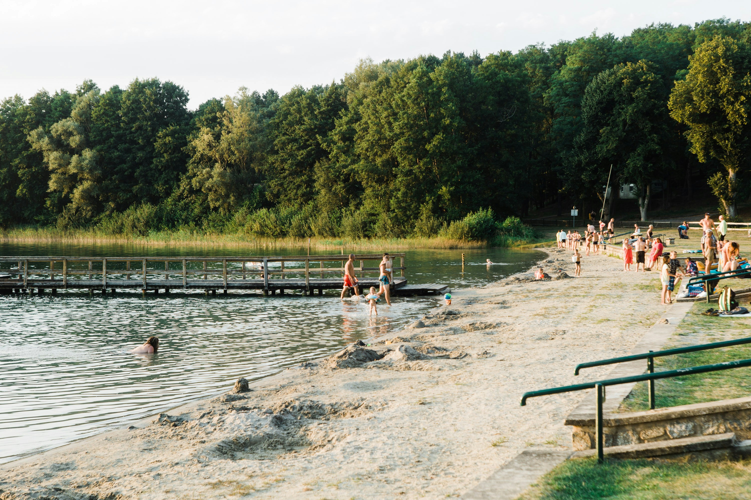 Families and friends enjoying a sunny day by the water, with a wooden dock and lush greenery in the background.