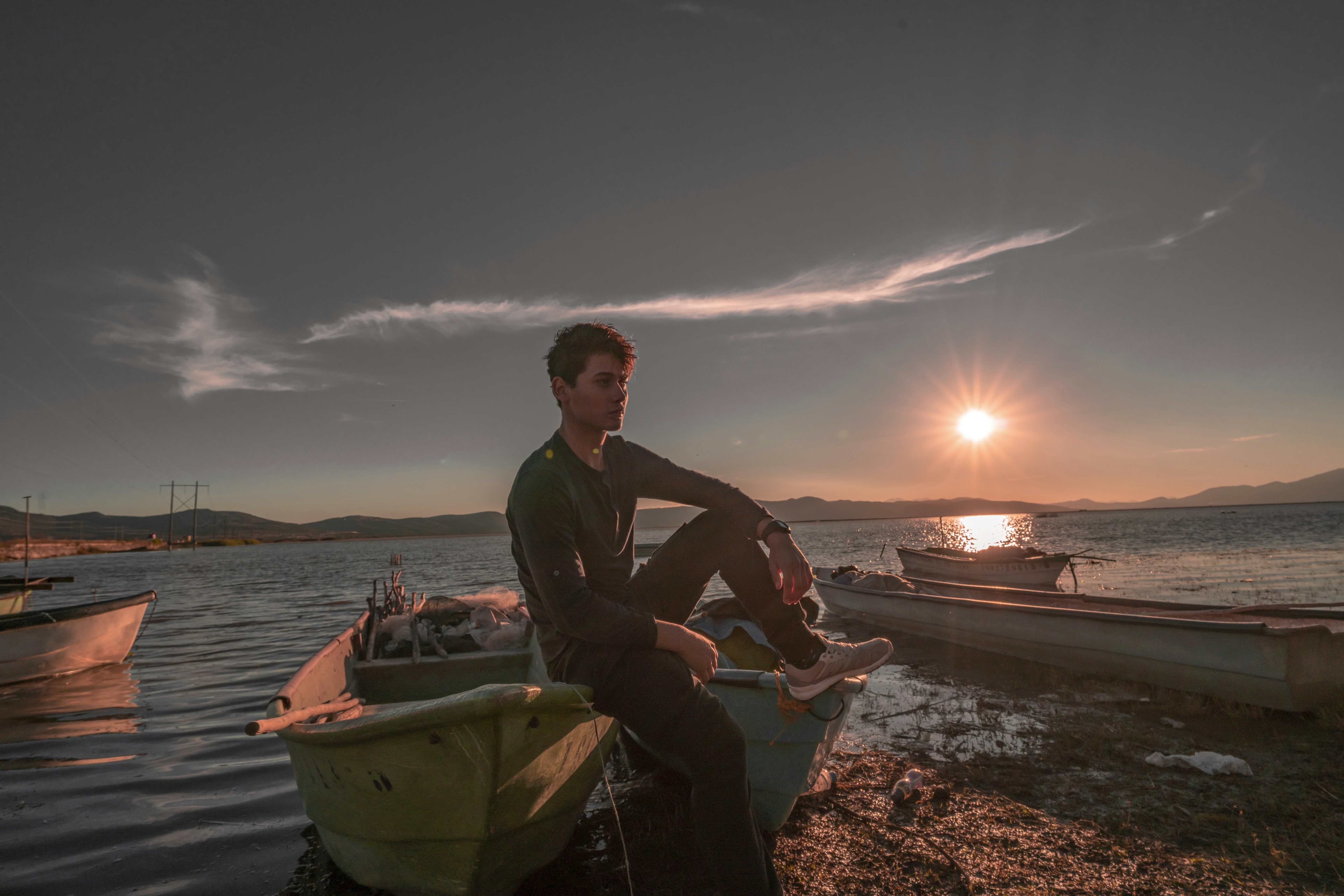 Young man sitting on the edge of a boat at sunset, surrounded by calm waters and distant mountains. The sun casts a warm glow over the scene.