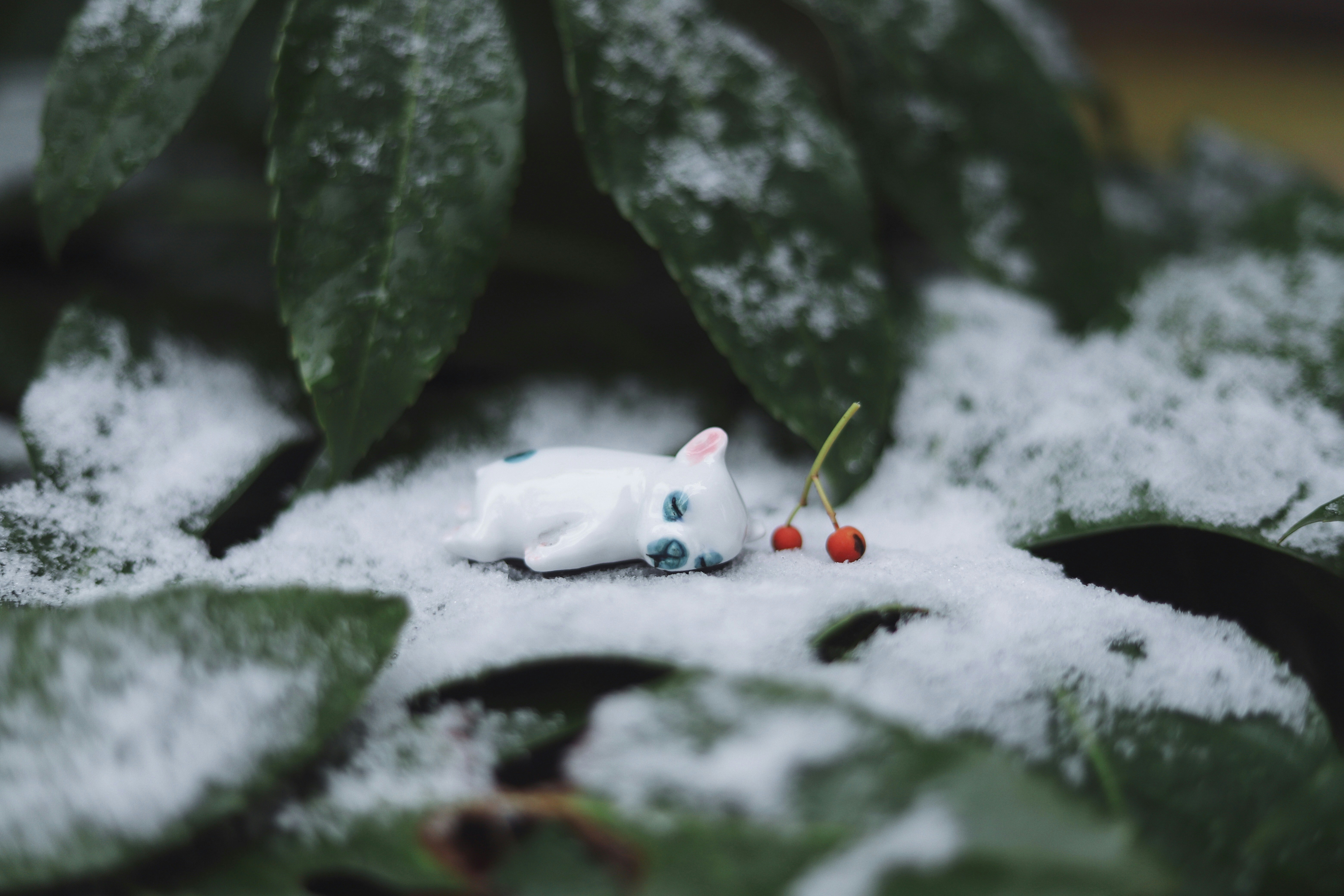A small white figurine rests on a bed of green leaves dusted with snow, accompanied by two bright red berries.