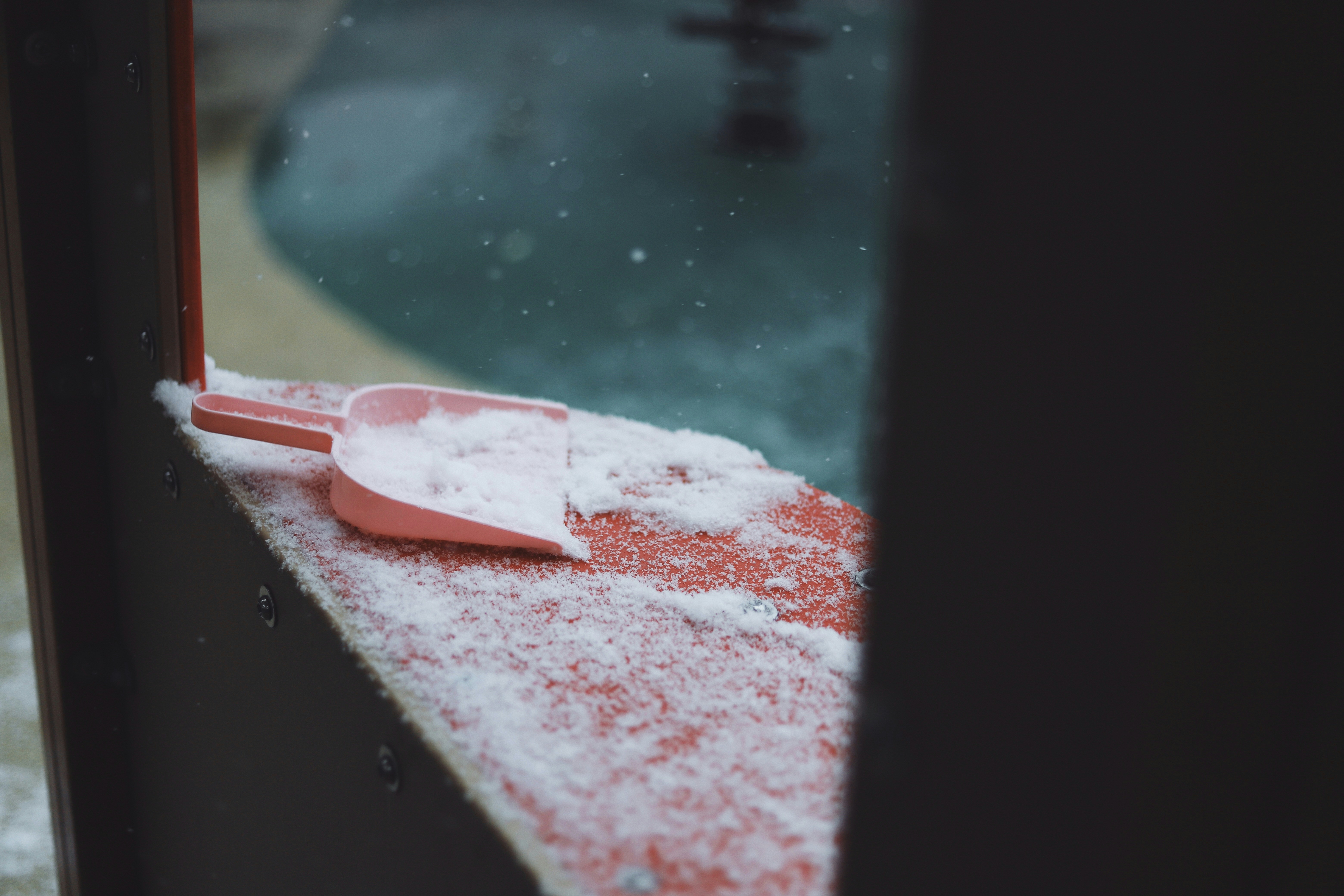 A pink snow shovel rests on a snow-dusted ledge, surrounded by a soft winter landscape. Flakes gently fall, adding to the serene atmosphere.