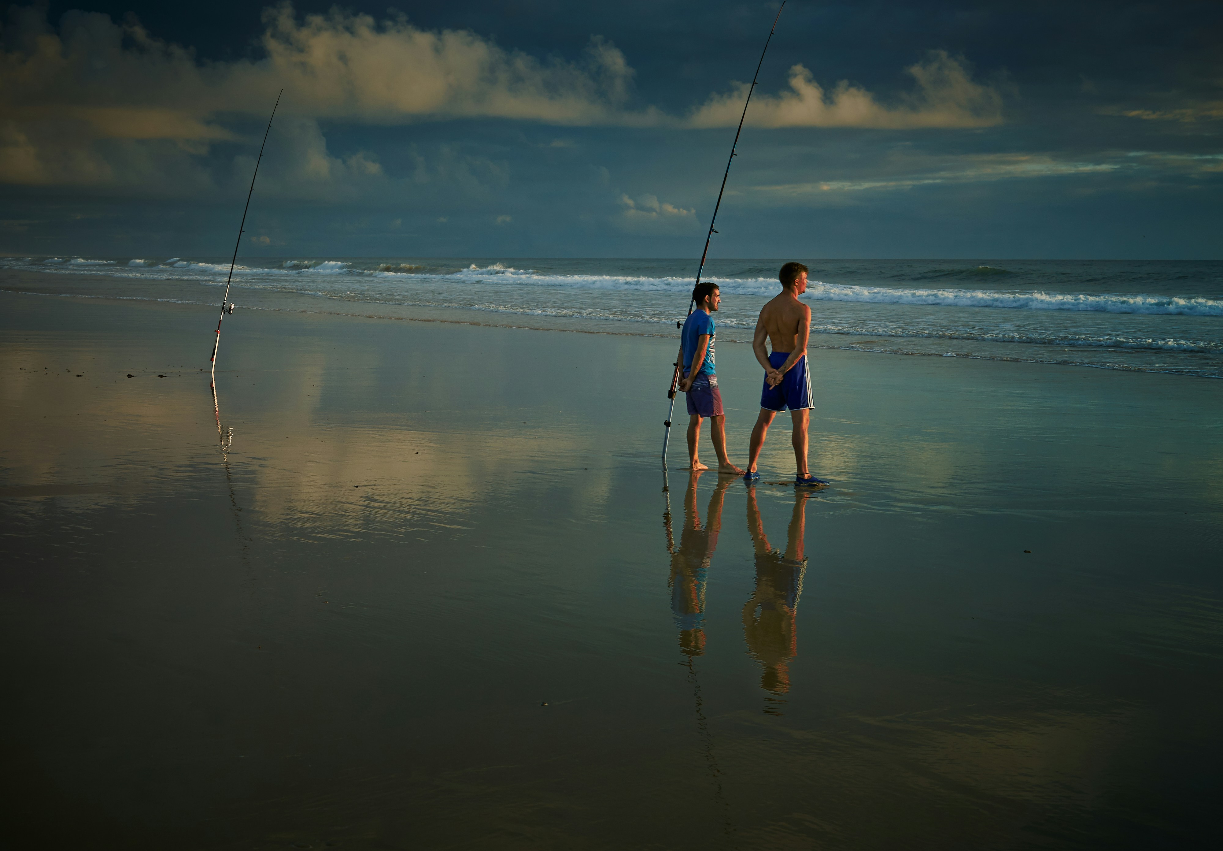 Two people fishing on a reflective beach under dramatic clouds.