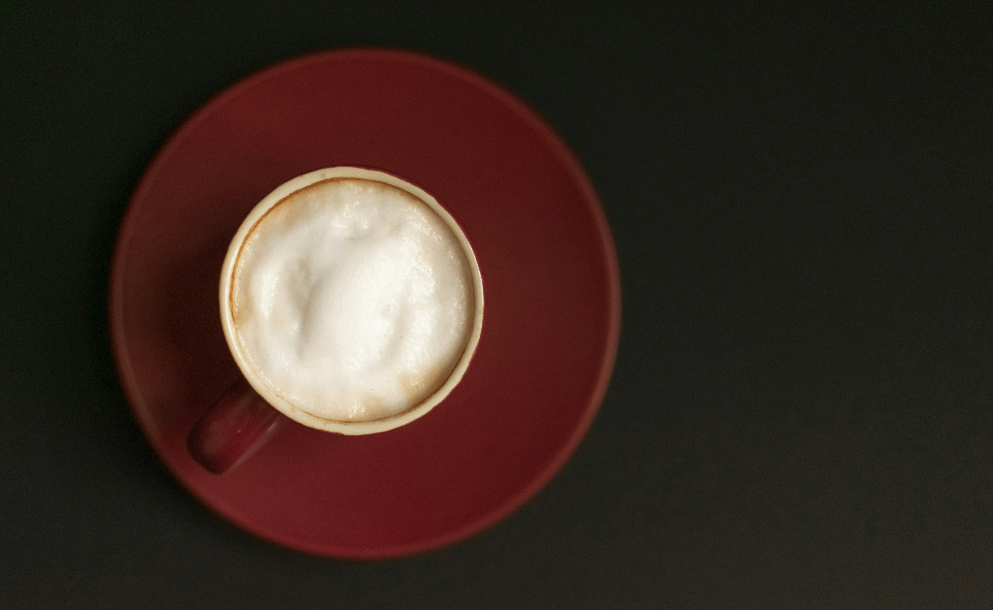 Creamy cappuccino topped with frothy milk sits on a deep red saucer against a dark background.