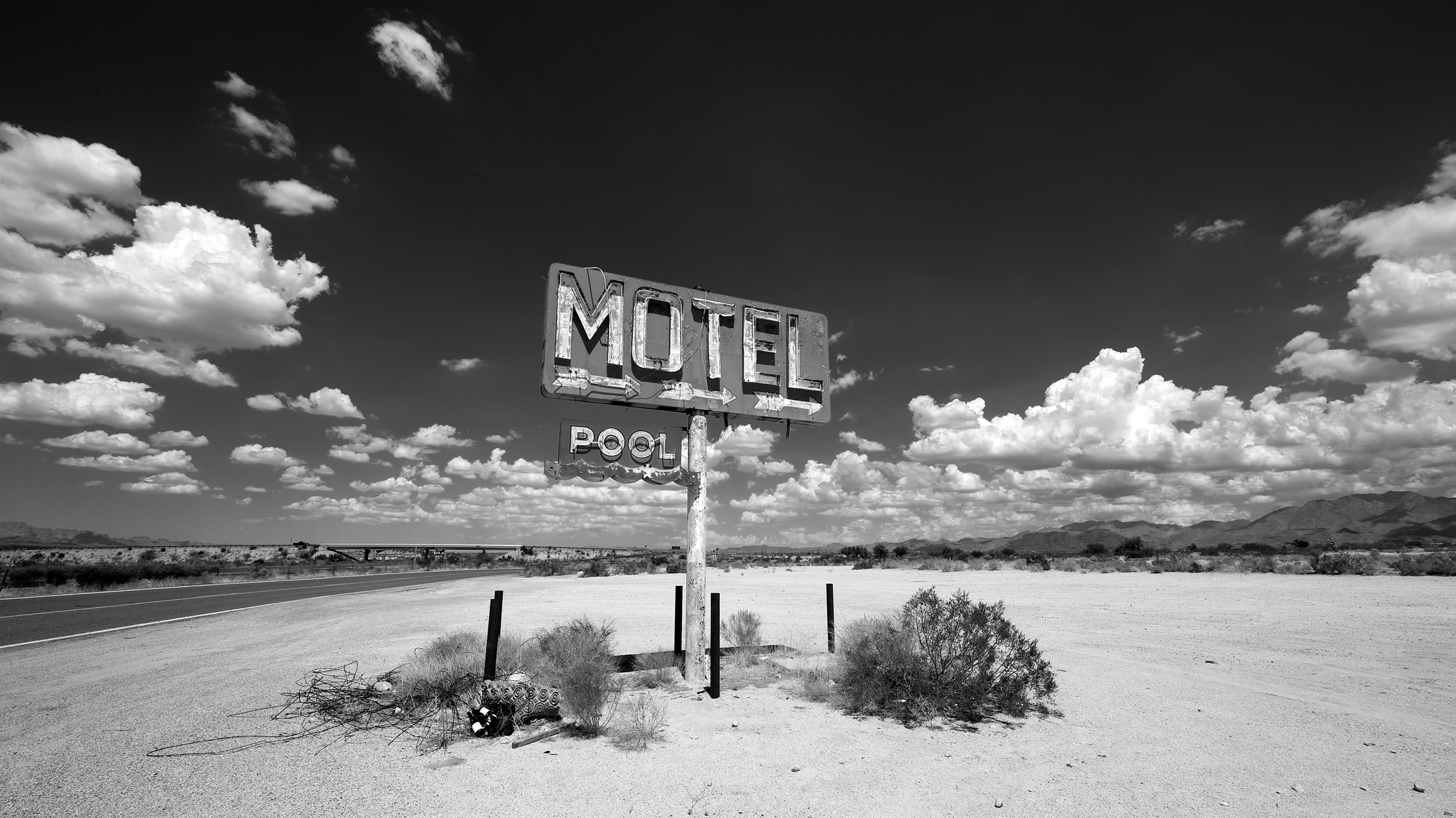 Weathered motel sign stands tall against a dramatic sky, surrounded by arid landscape and scattered vegetation.