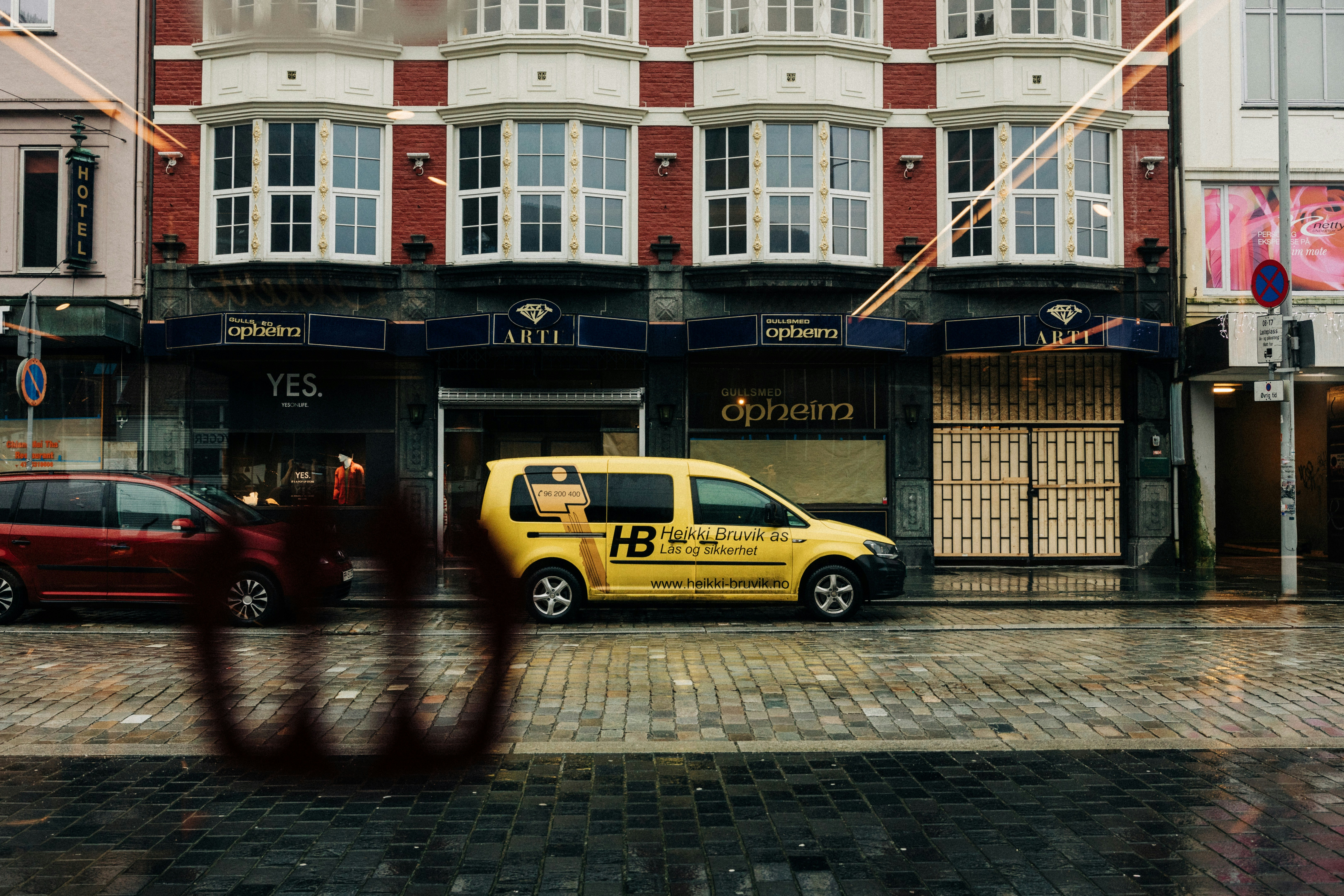Yellow delivery van parked on a rainy street in an urban setting, with reflections visible on wet pavement and buildings in the background.