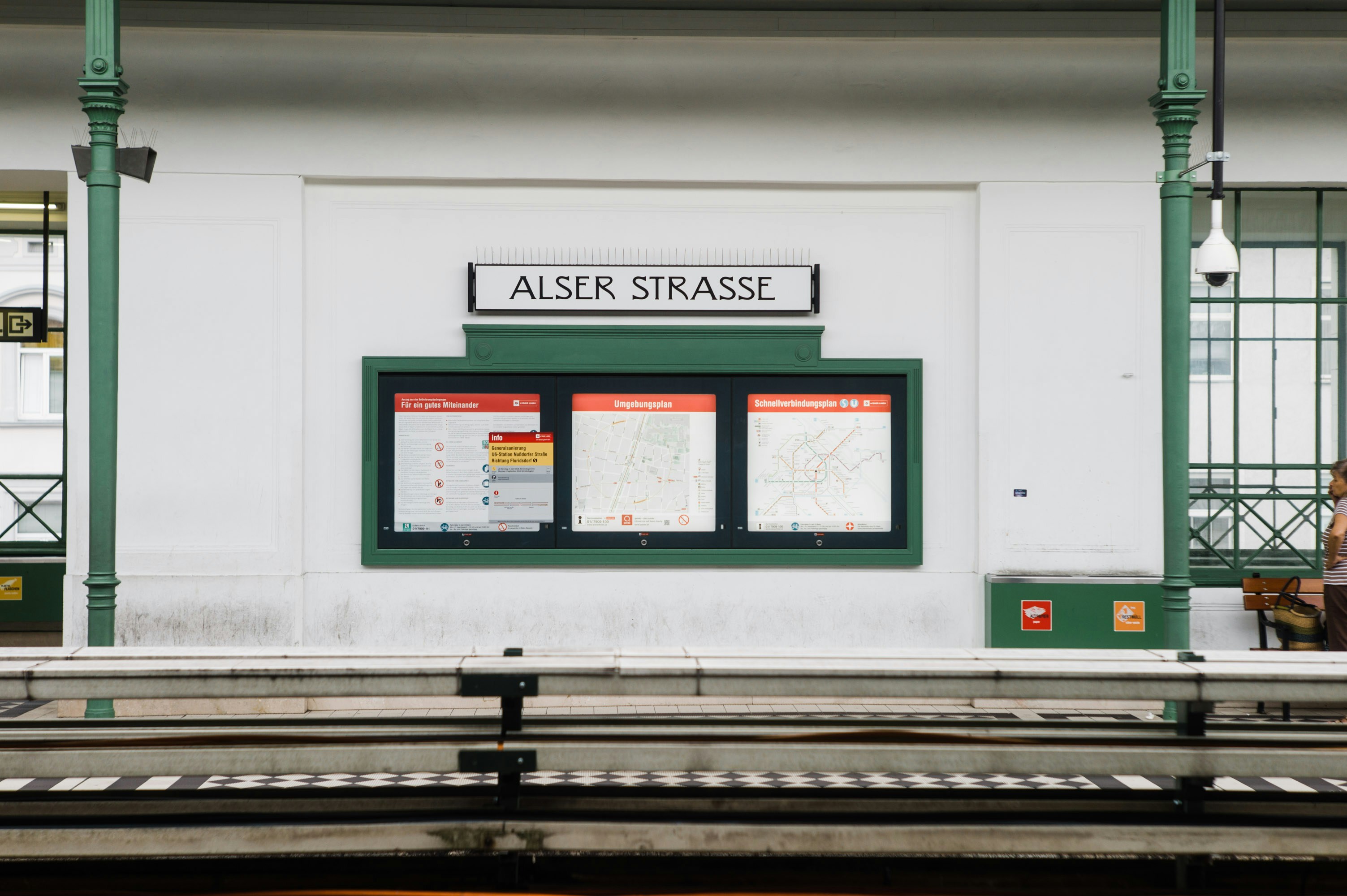 Station wall featuring the 'ALSER STRASSE' sign above three framed map panels on a white facade, with green trim and platform rails in the foreground.