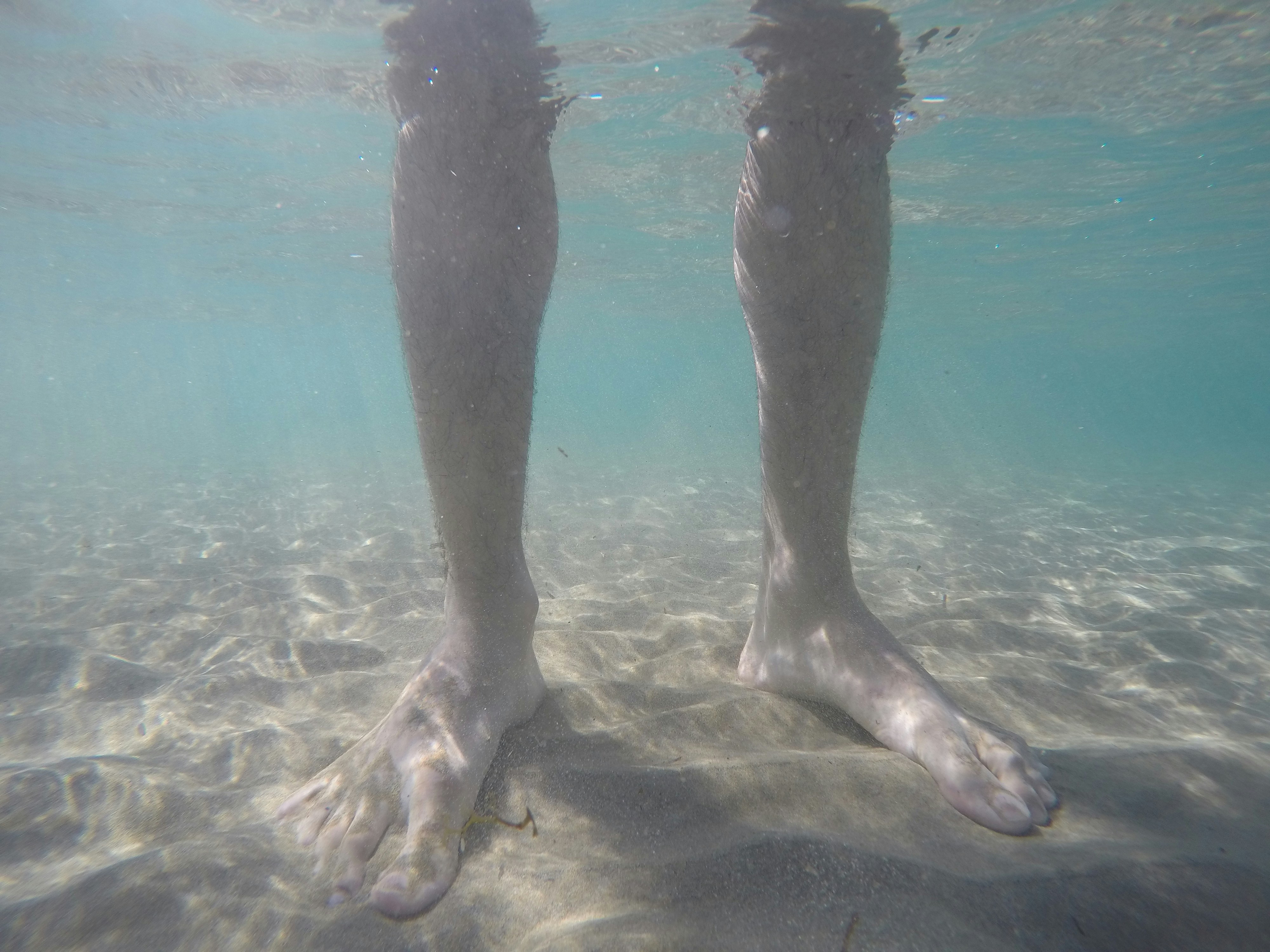 Underwater photograph of two legs standing in shallow, sunlit lagoon sand, with feet submerged and rippling light patterns.