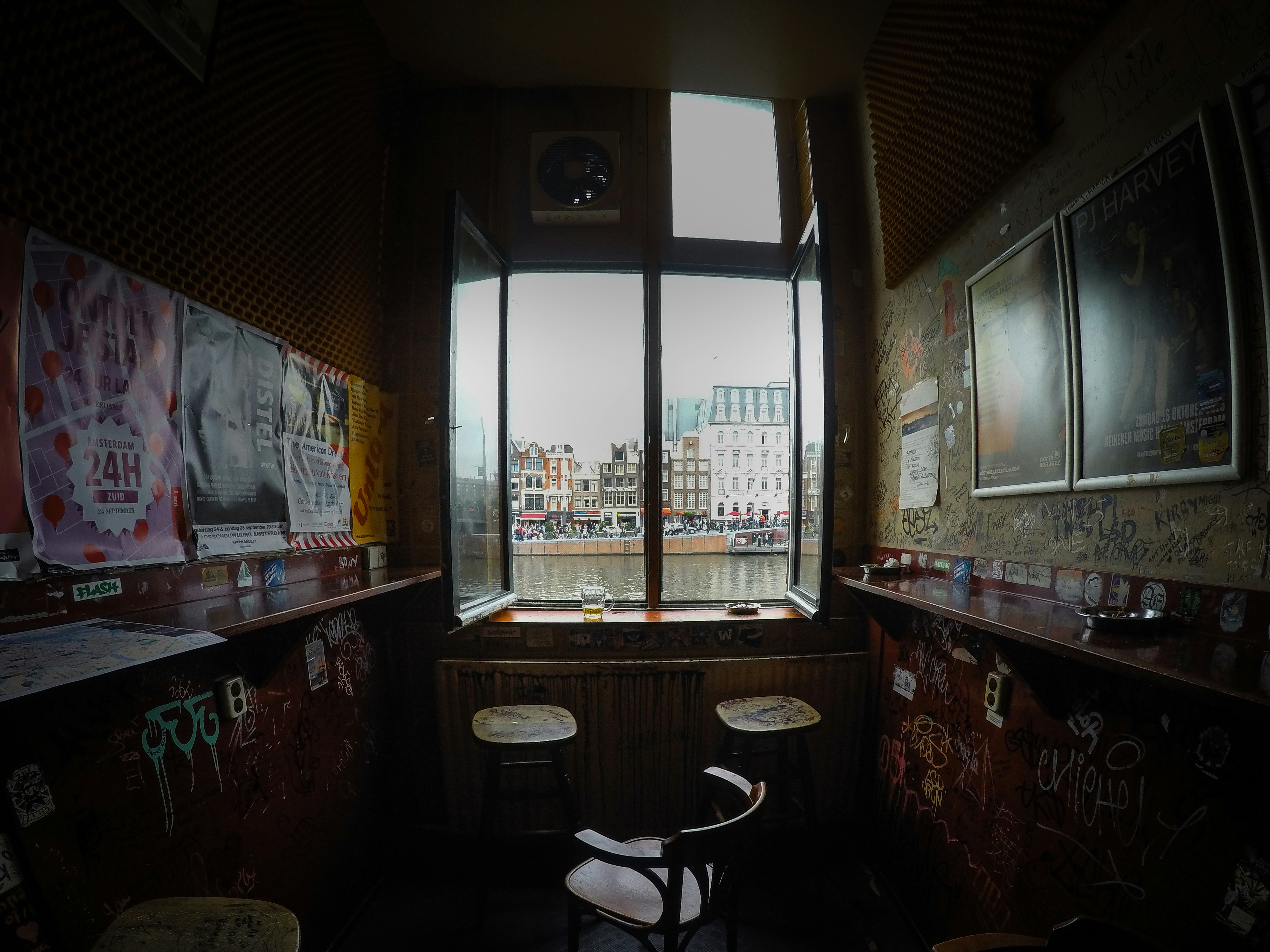 Graffiti-covered barroom interior with a large window framing a distant city canal and historic buildings, flanked by two stools and poster-covered walls.