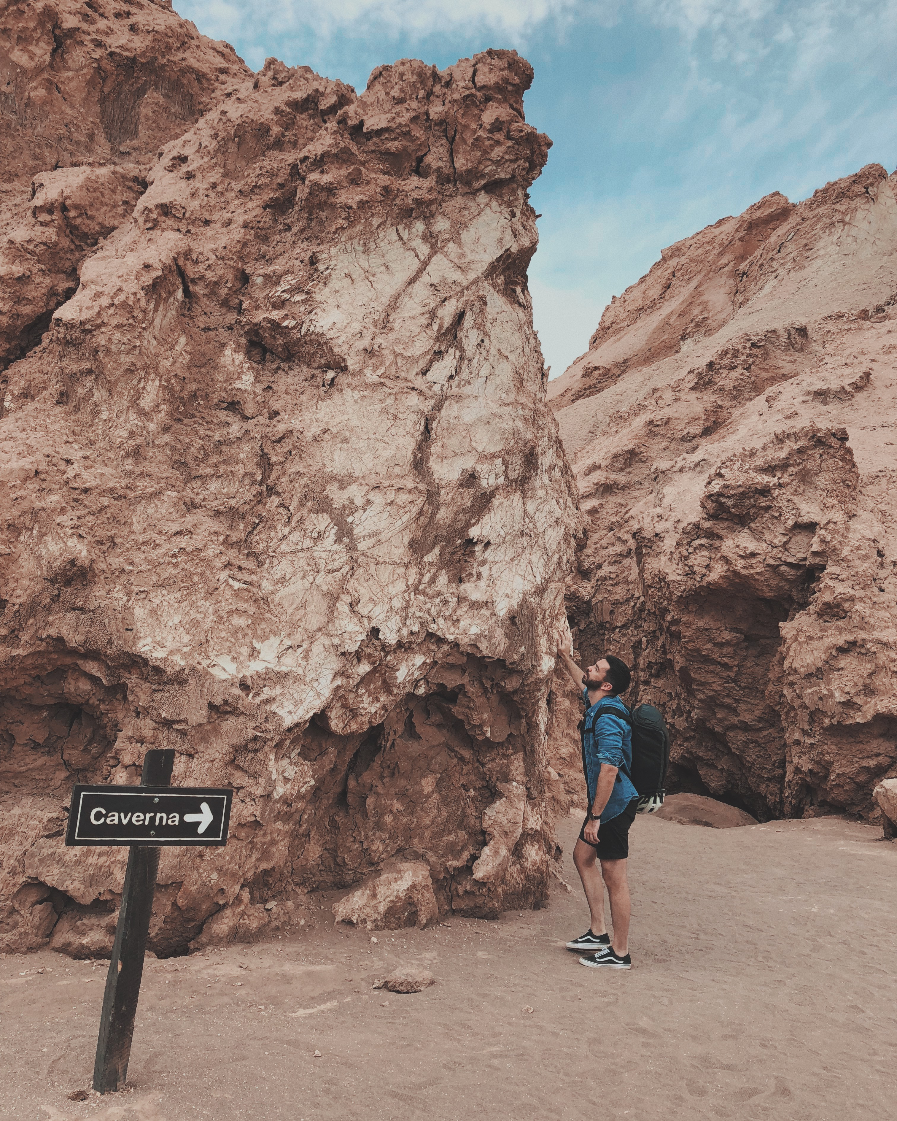 A traveler gazes up at a towering rock formation, with a directional sign pointing towards a cave entrance. The rugged landscape showcases natural erosion patterns.