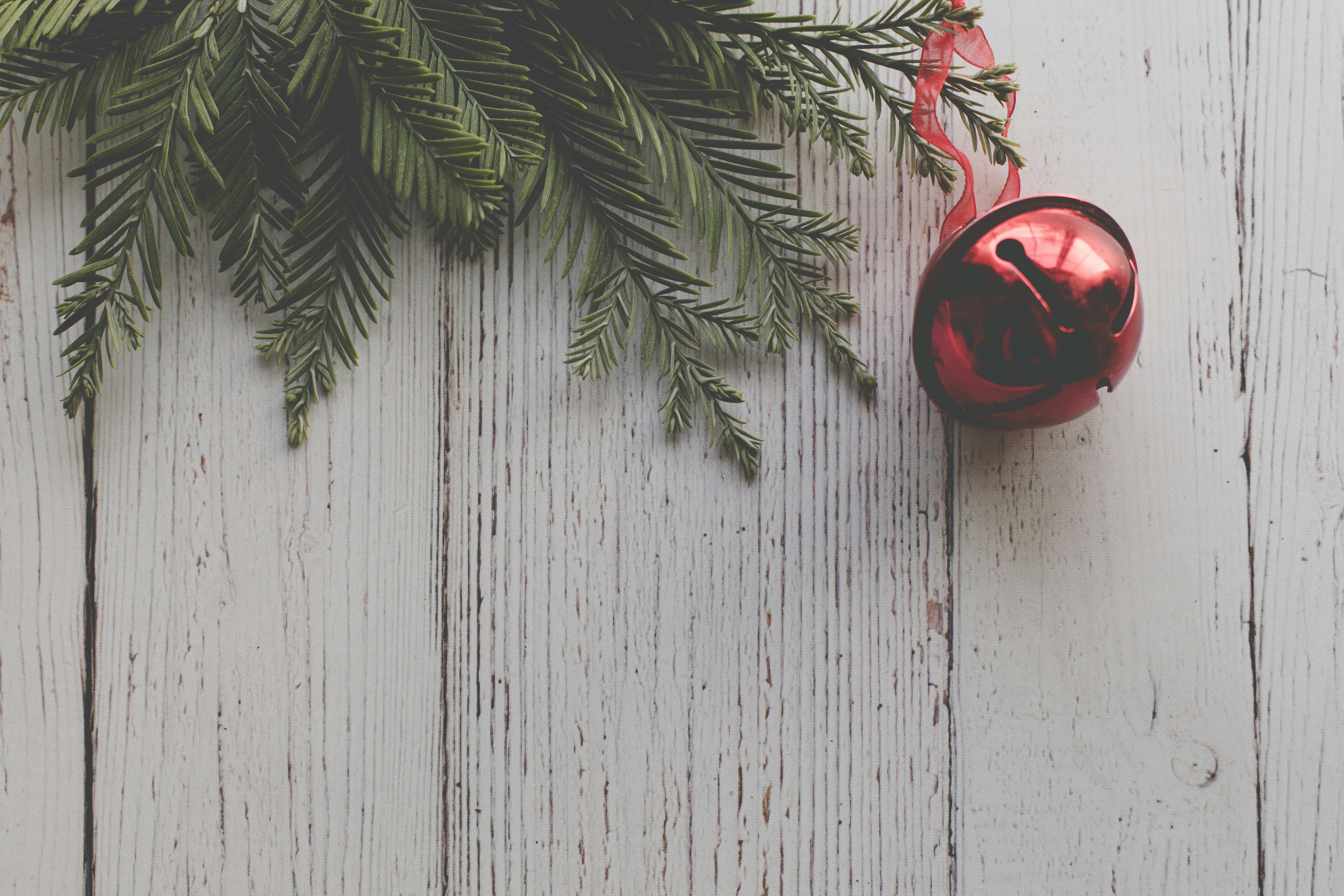 A vibrant red bell adorned with a ribbon rests beside lush green pine branches on a weathered white wooden surface.
