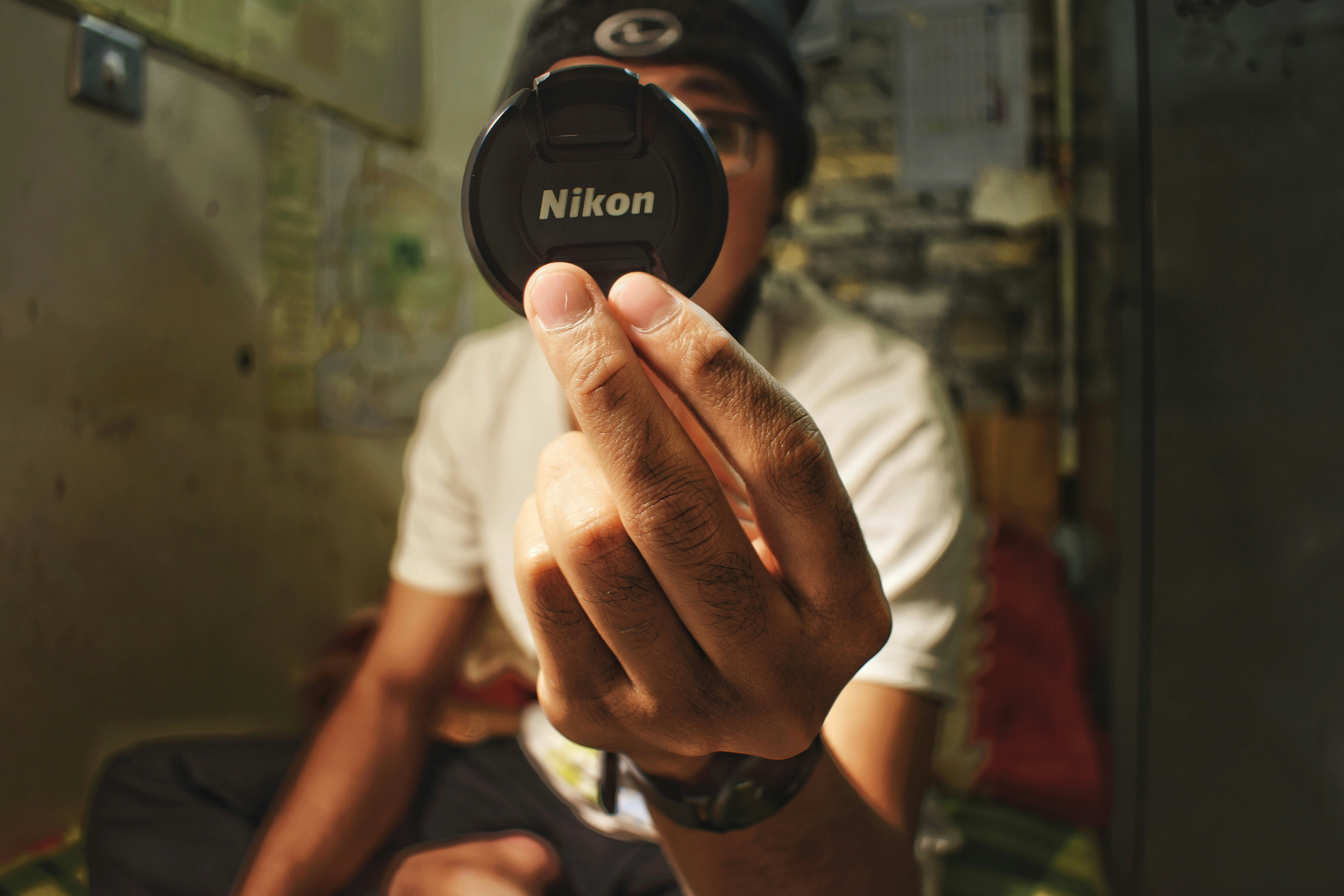 Person holding a Nikon lens cap in a dimly lit room, with a blurred background.