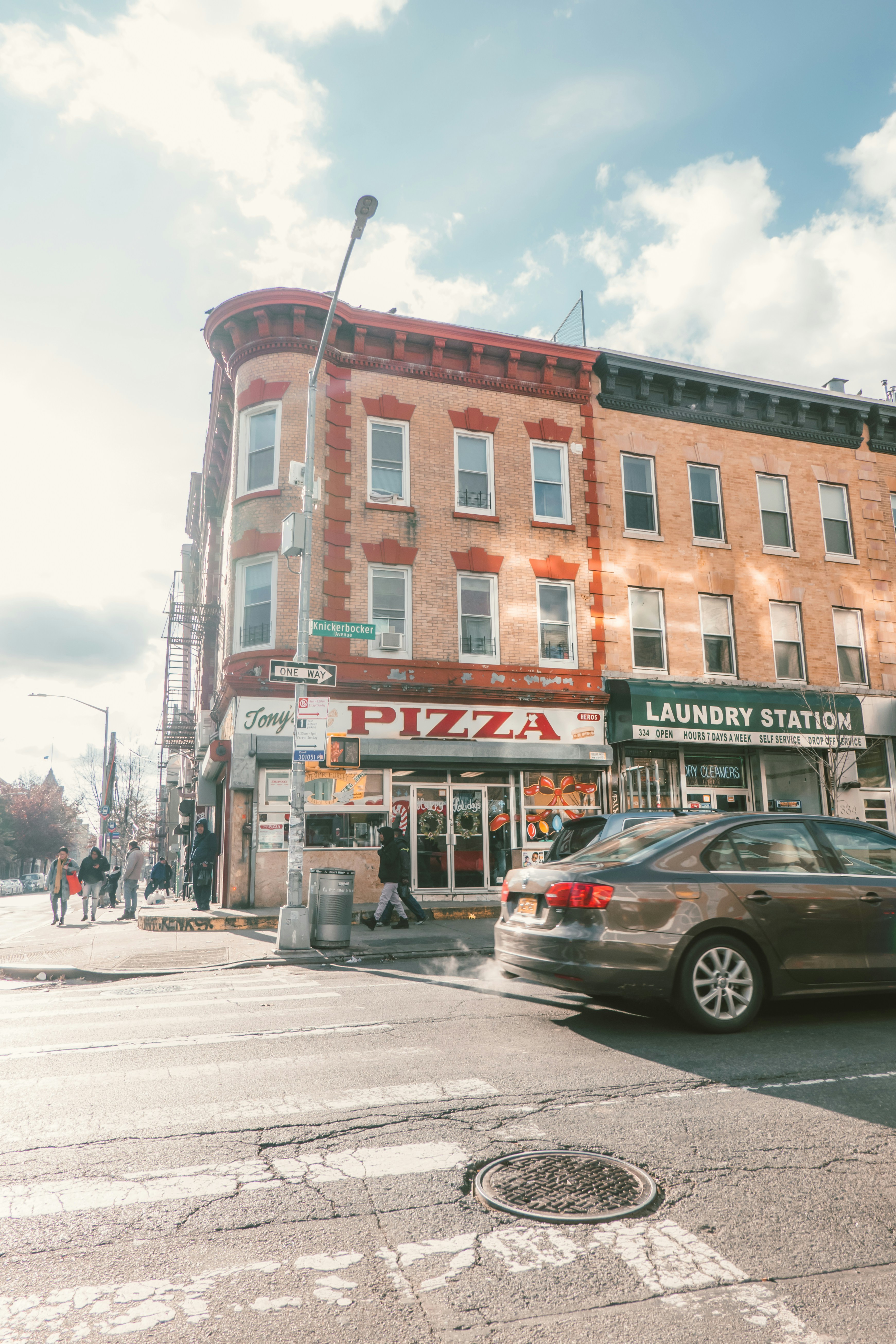Tony's Pizza storefront bustling with pedestrians and a car passing by on a sunny day. Laundry Station sign adds to the urban vibe.