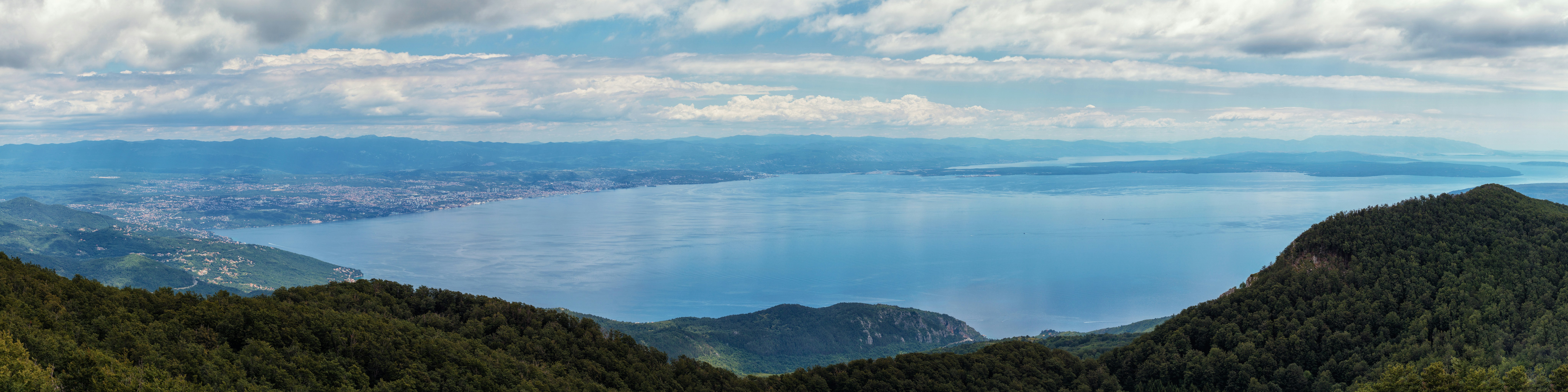 Expansive view of a coastal city bordered by mountains and a vast sea under a partly cloudy sky.