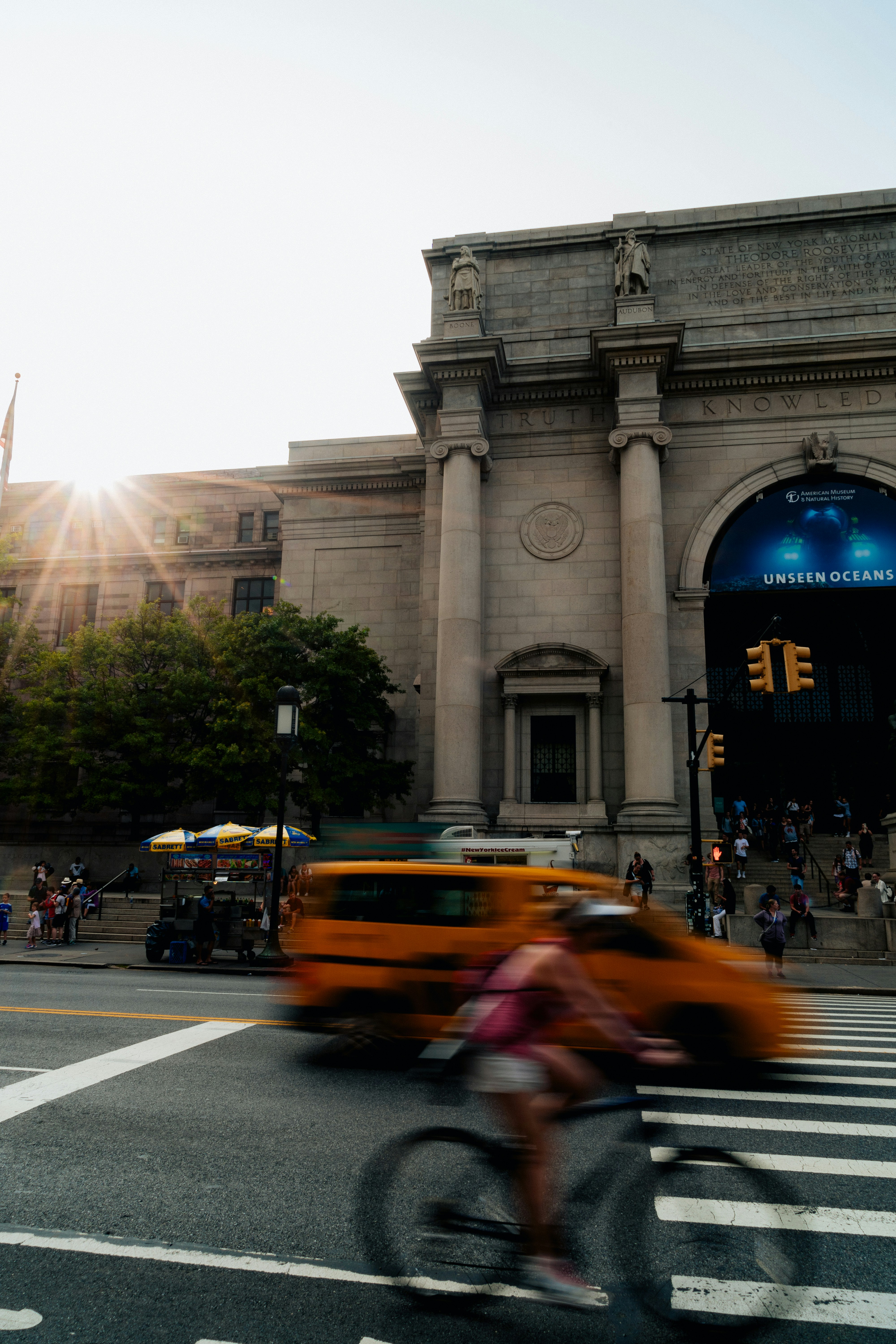 Blurred cyclist and taxi passing a grand stone building as the sun sets.