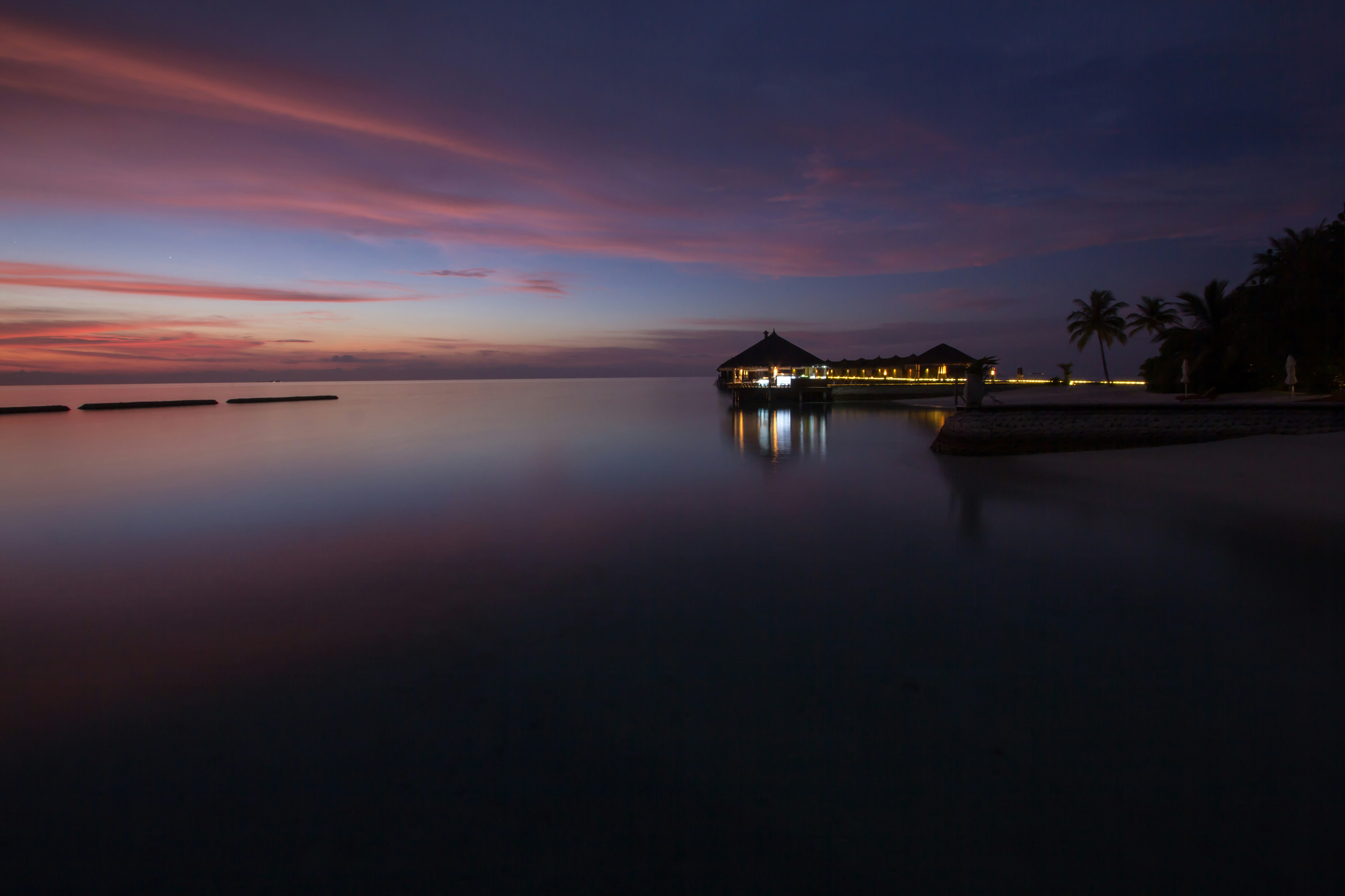 Serene ocean view with a distant lit pavilion and palm trees under a twilight sky with pink and purple hues.
