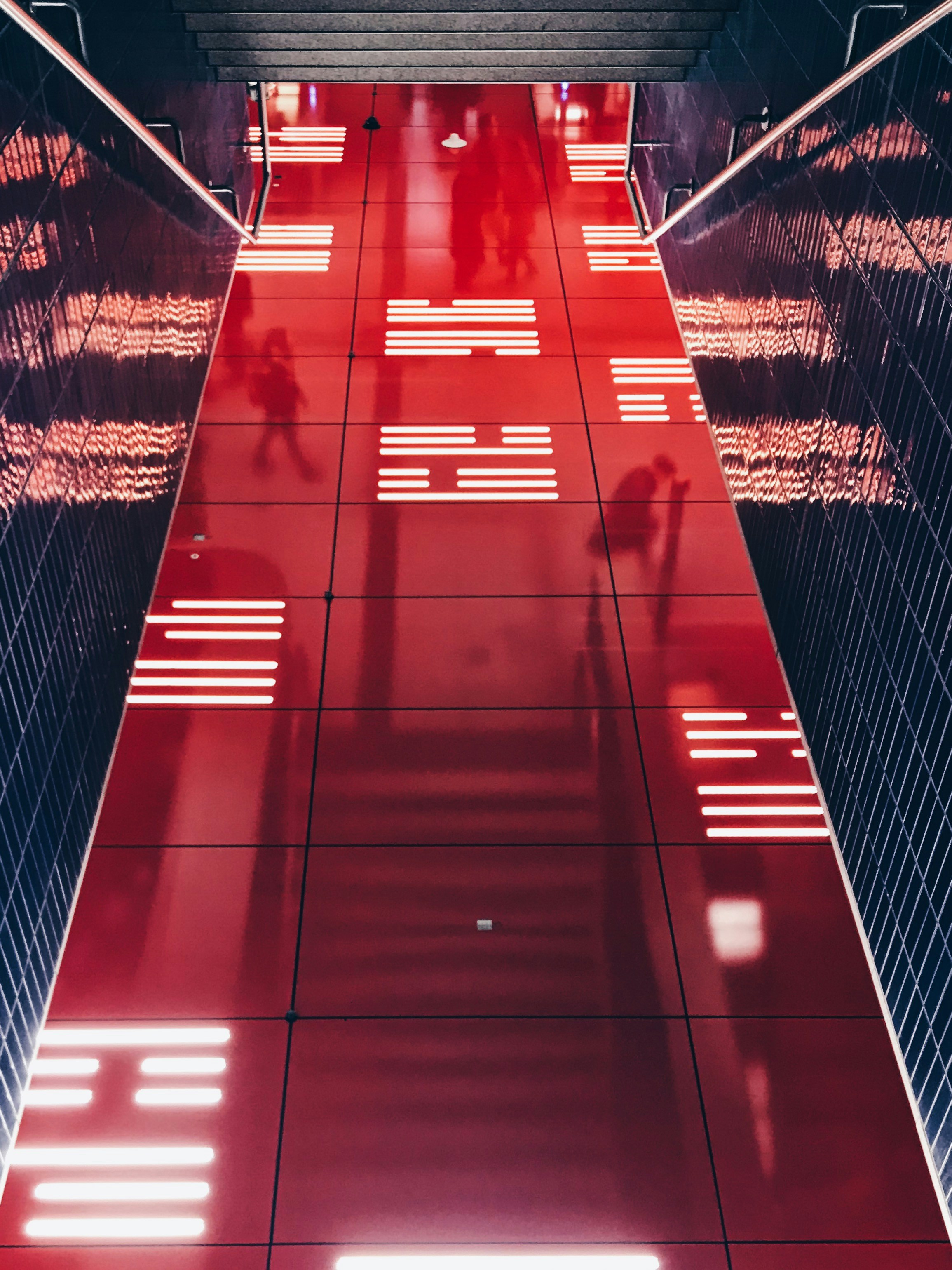 Vibrant red tiled floor illuminated by overhead lights, reflecting shadows of people descending stairs.