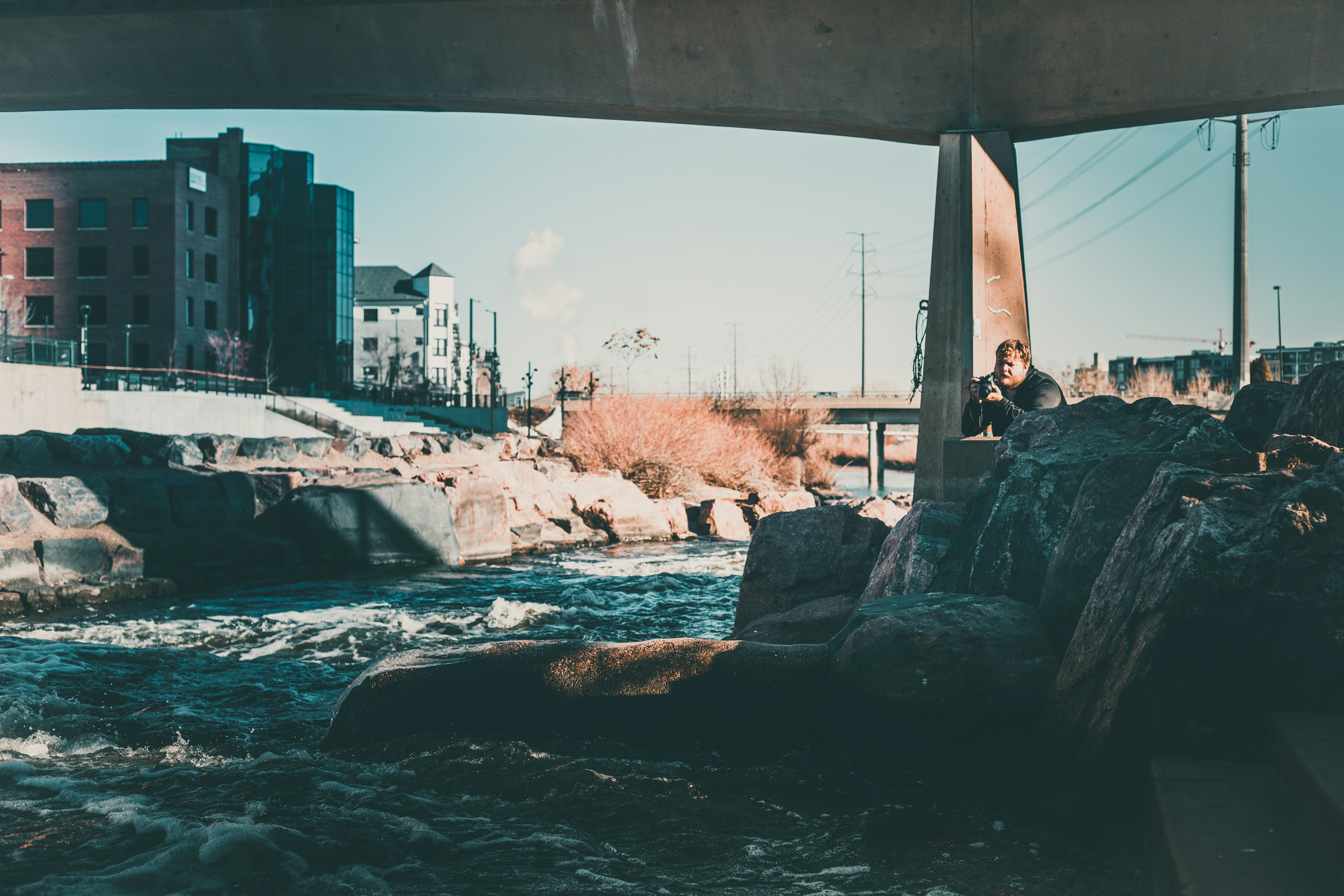 A person sits on large rocks by a flowing river, framed by a concrete overpass and urban buildings in the background. The scene captures a moment of tranquility amidst city life.