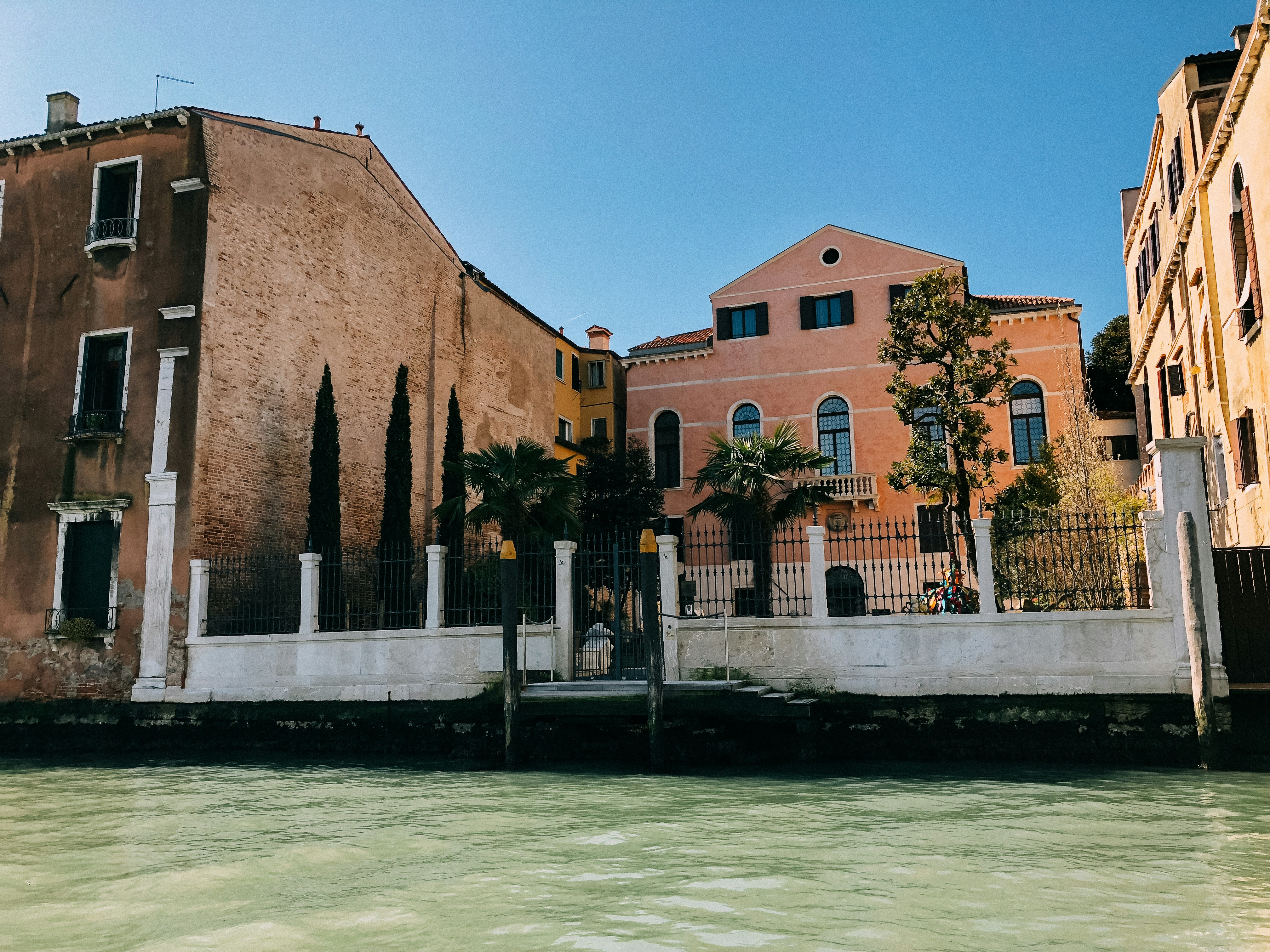 Sunlit Venetian buildings and trees reflected in calm canal waters under a clear blue sky.
