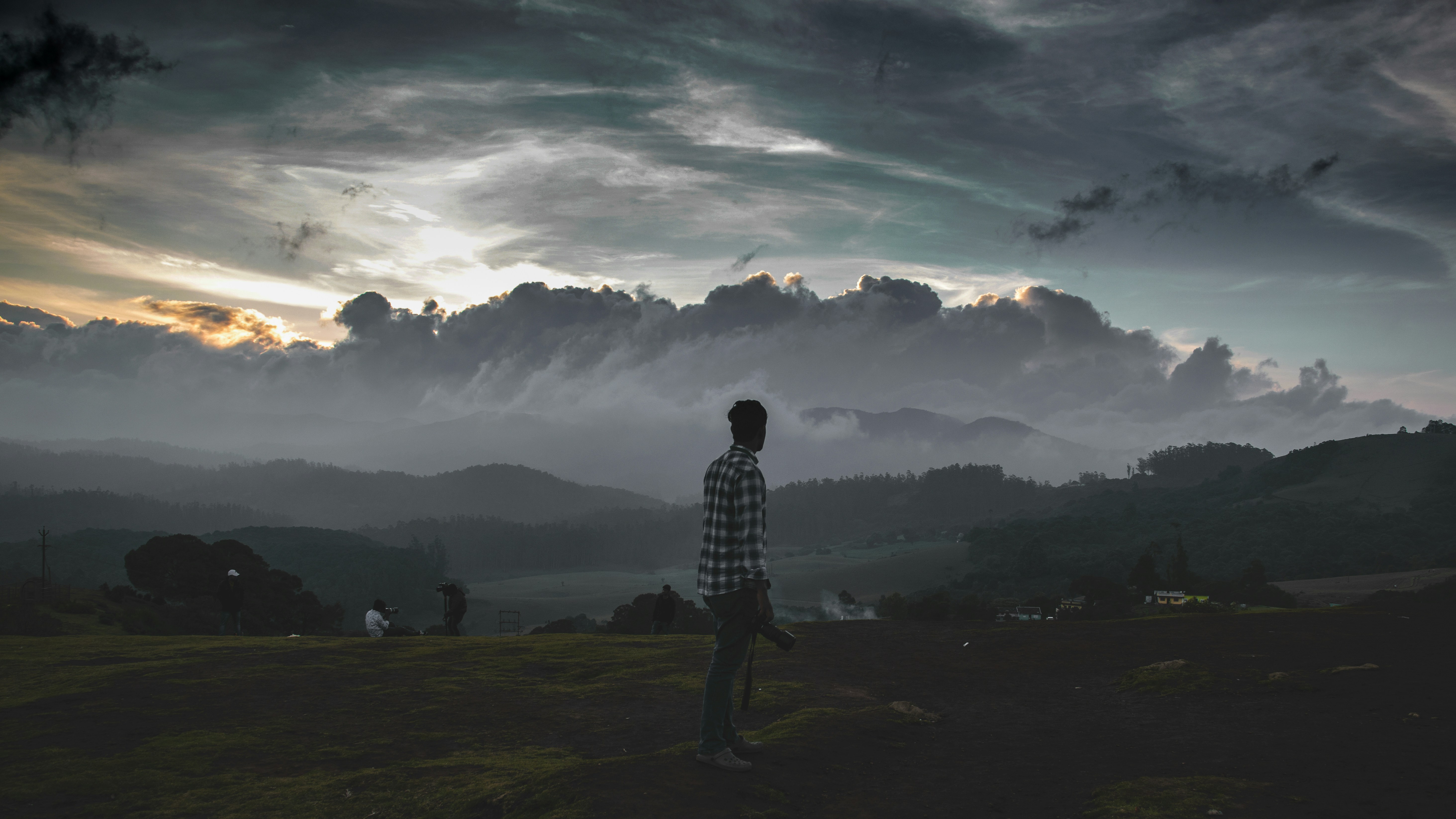Silhouette of a person standing against a dramatic sky filled with clouds and distant hills at twilight.