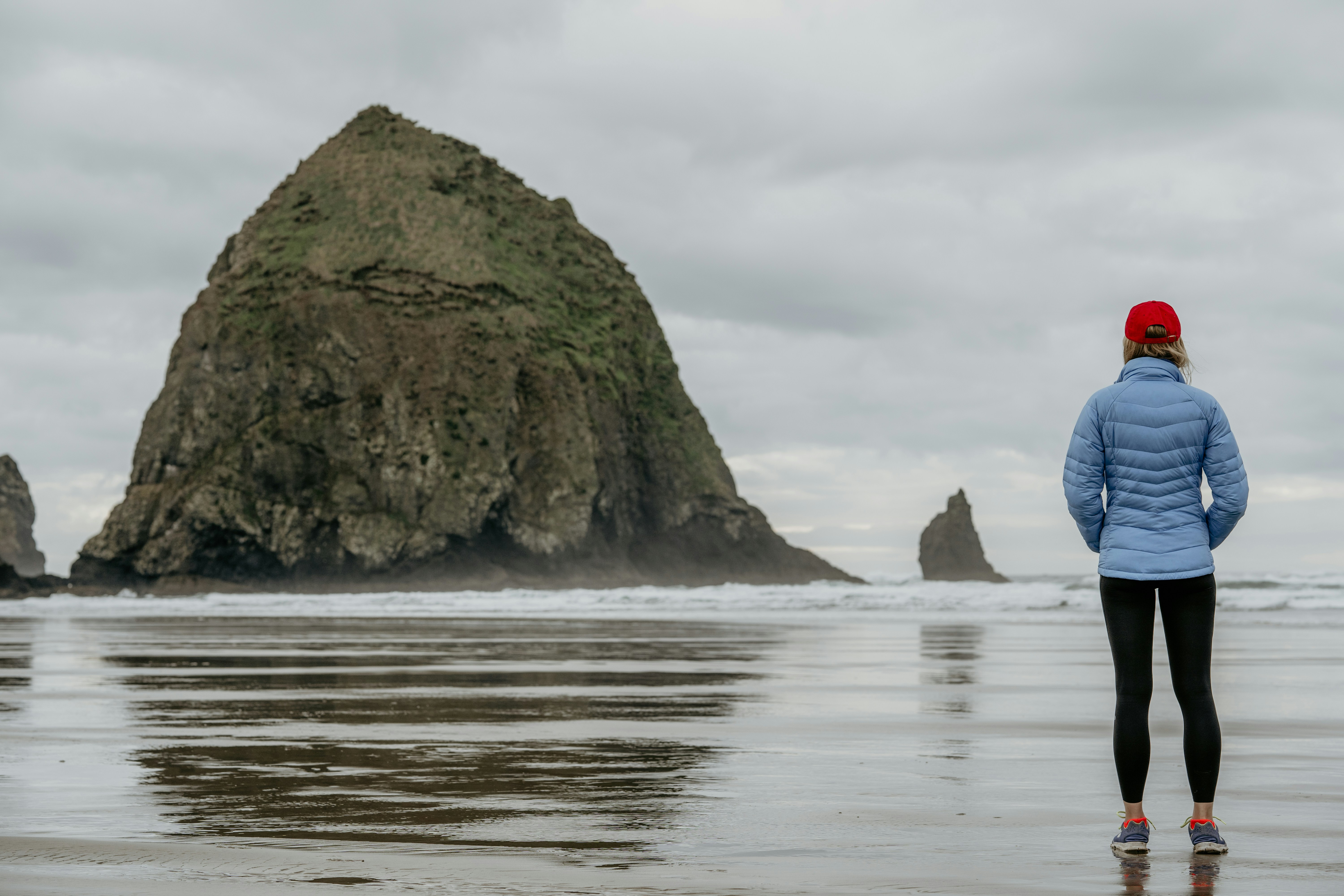 Red-Capped Gaze: A Lone Figure Framed by Sea Stacks on a Misty Beach