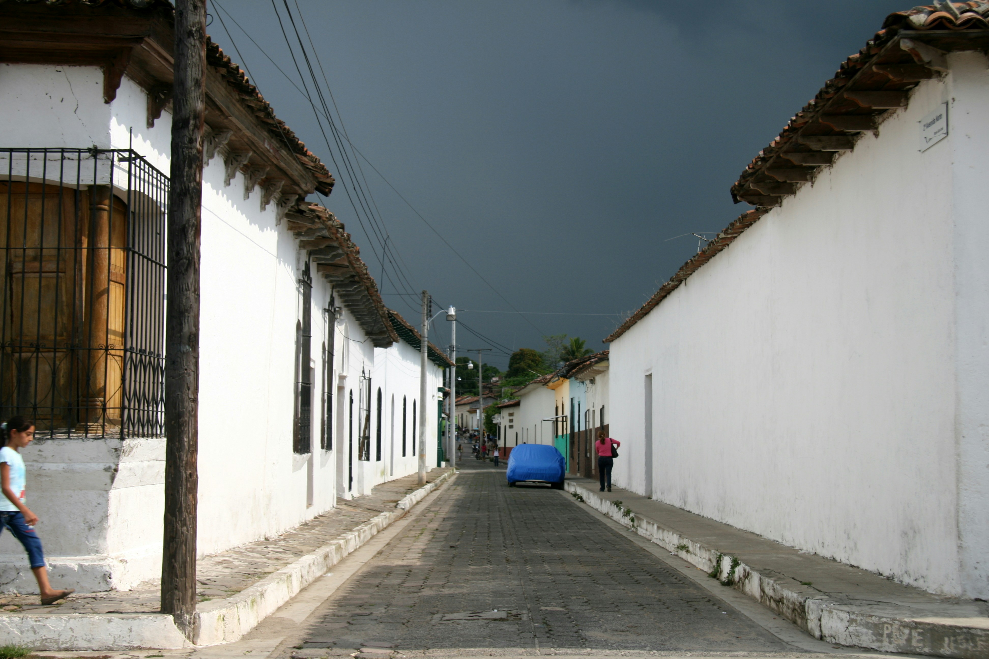 Narrow street lined with whitewashed buildings and a blue car, contrasting against an ominous dark sky. A woman walks past, adding a sense of life to the scene.