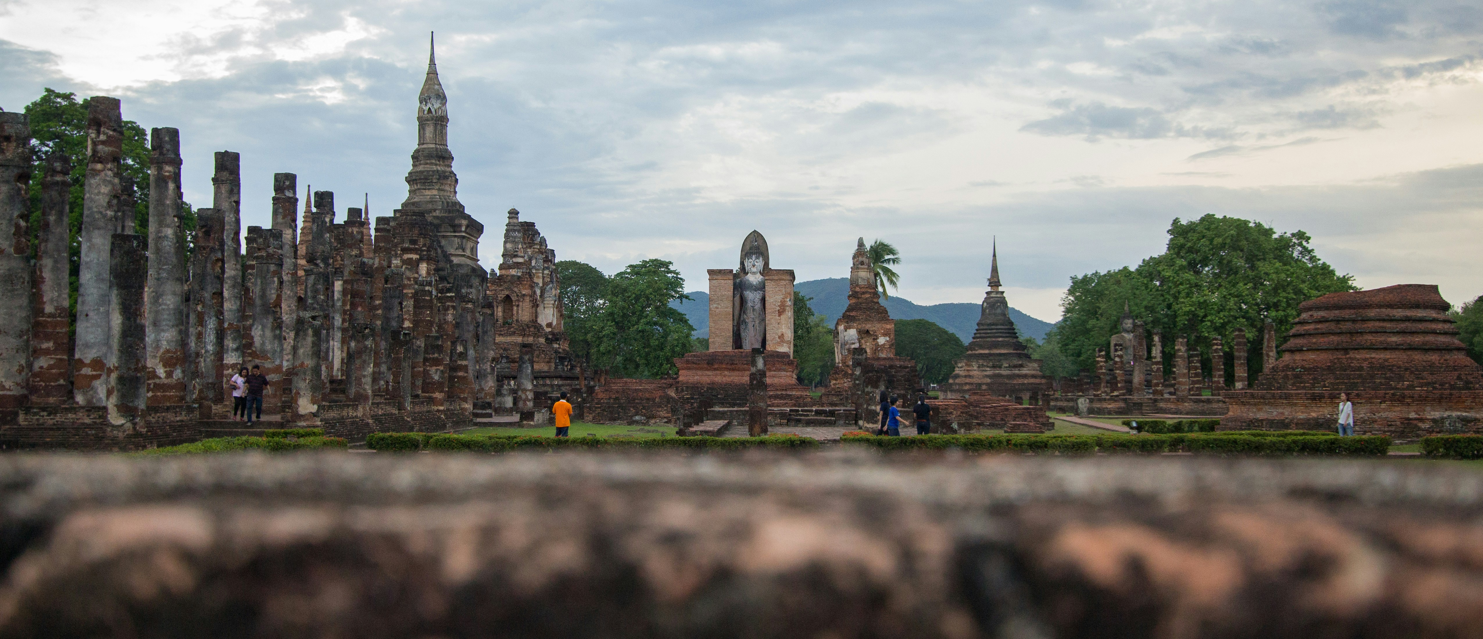 Ruins of a historical site with crumbling structures and trees, set against a cloudy sky. Visitors explore the remnants of a once-great civilization.